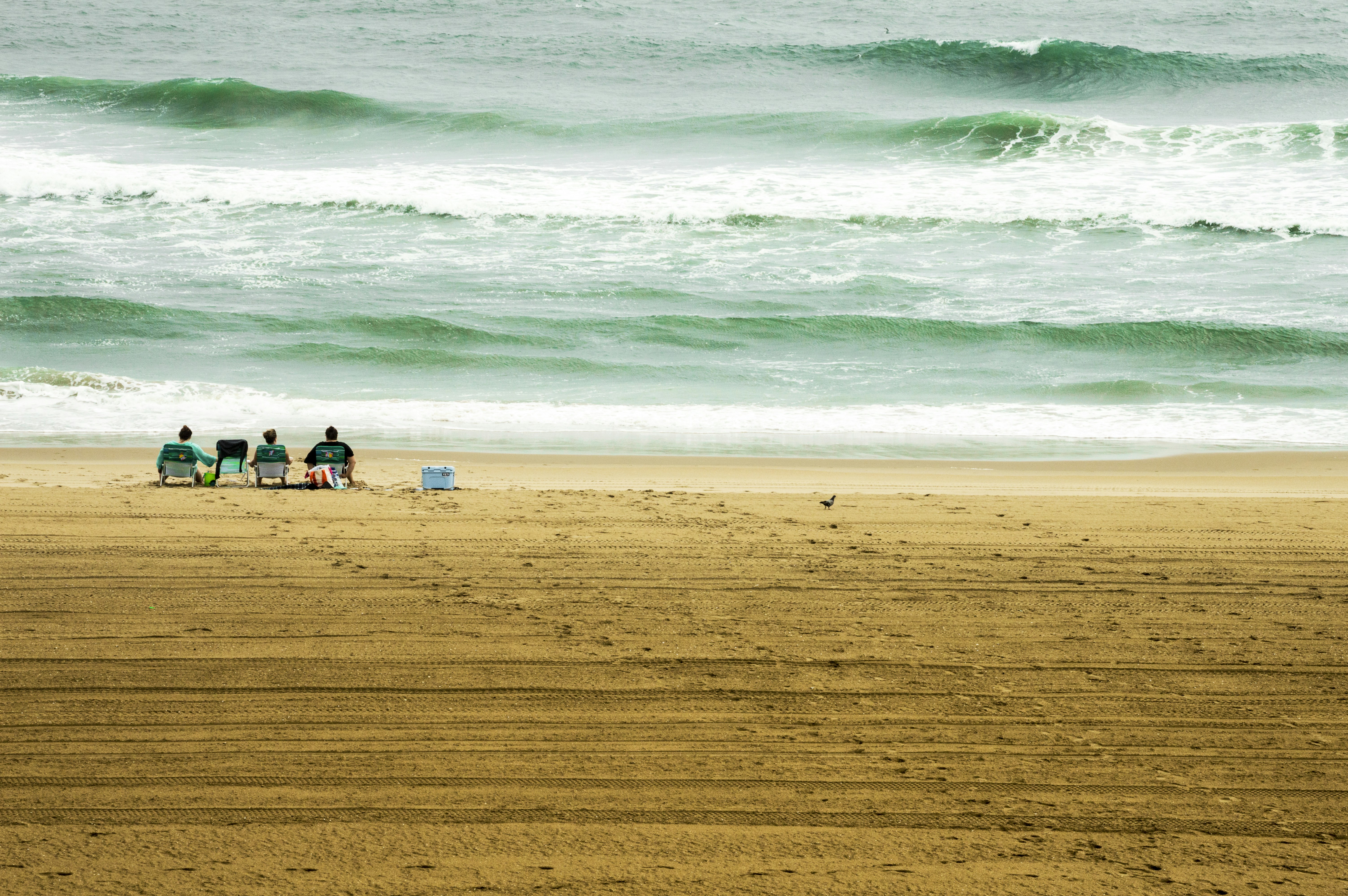 Three people seated on a sandy beach facing rolling ocean waves.