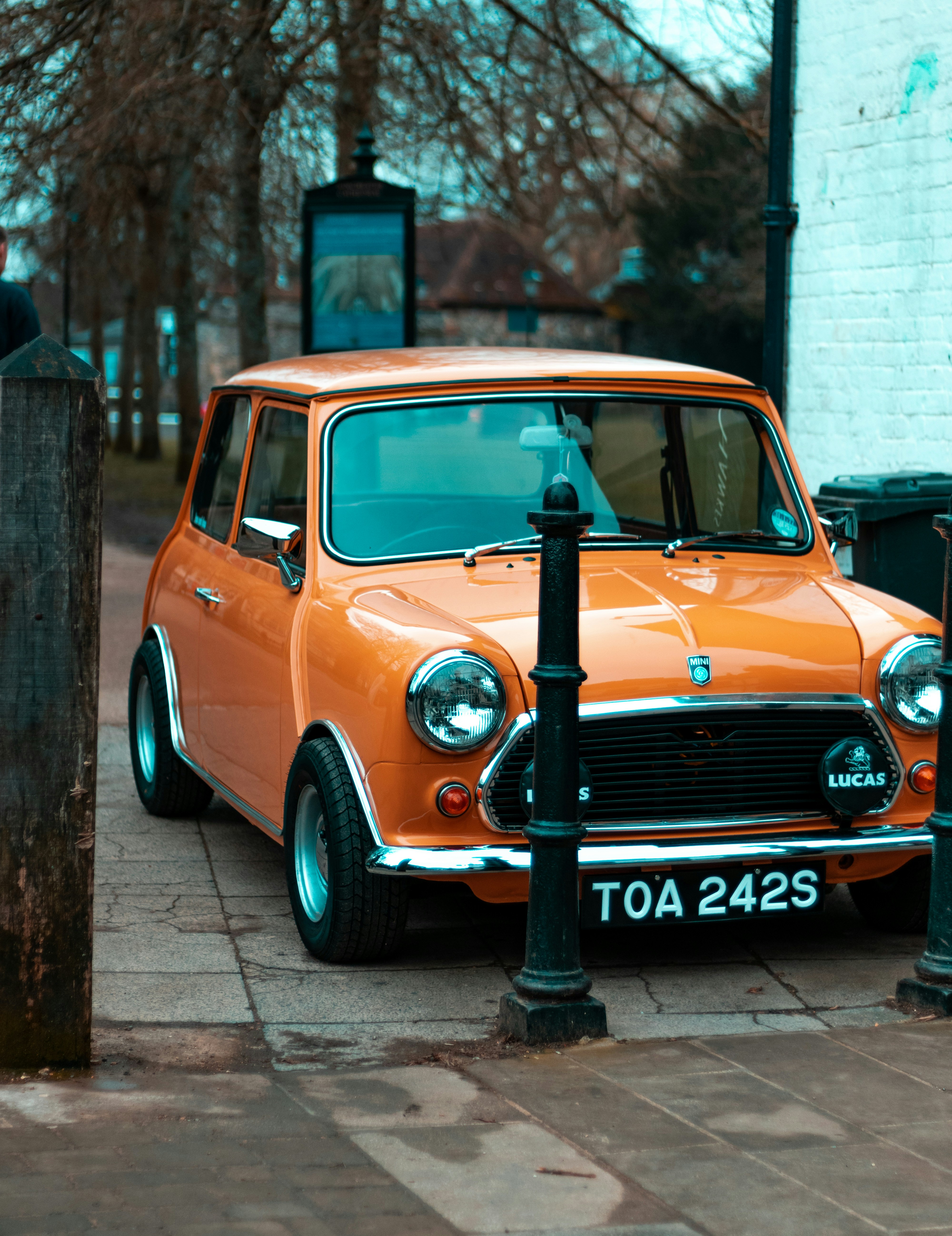 Bright orange vintage car parked beside a lamppost in an urban setting, surrounded by trees and buildings.