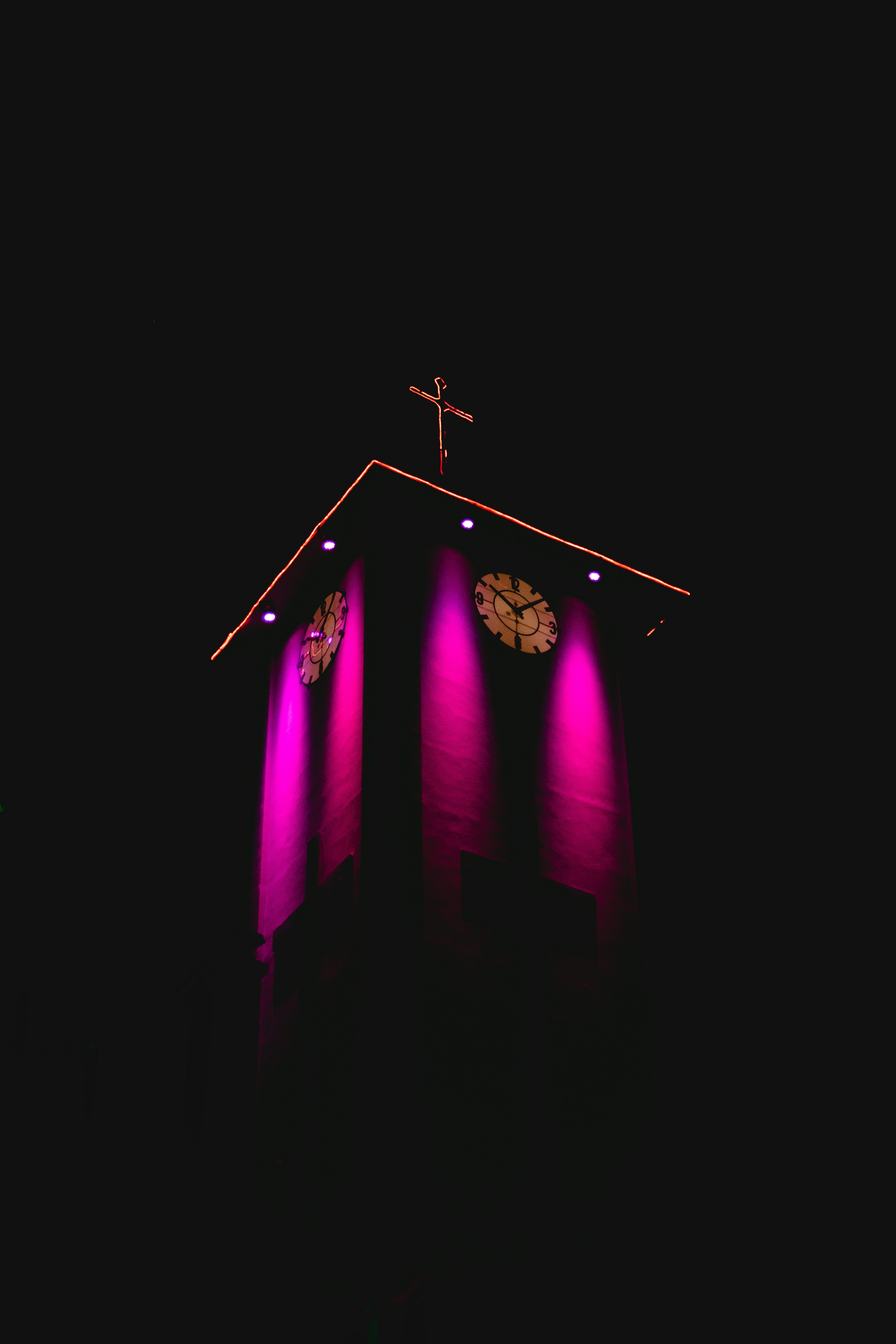 A church tower illuminated with vibrant pink lights, featuring a cross at the top and two clock faces. The scene captures a blend of spirituality and modern aesthetics.