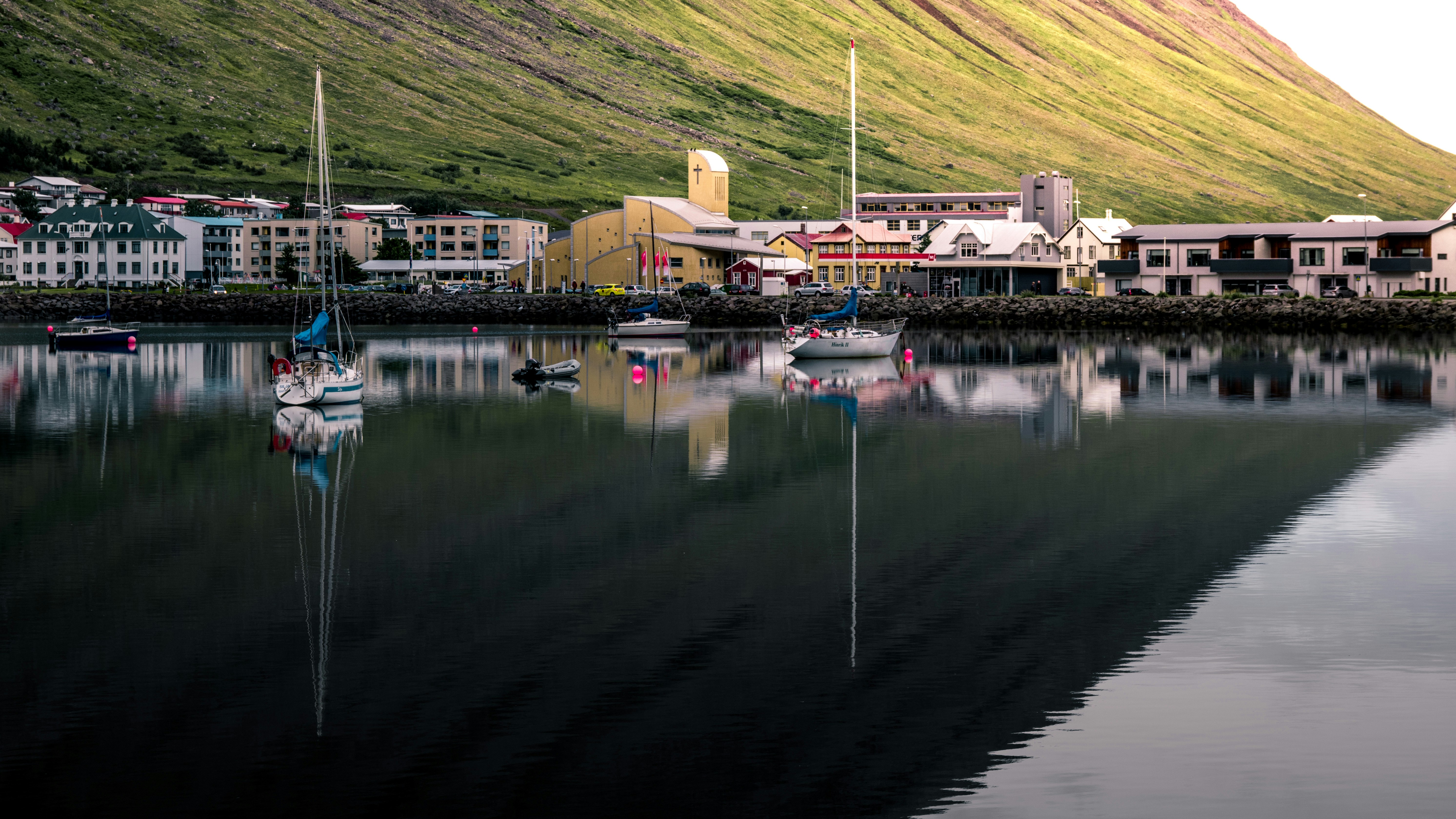 Colorful boats anchored in a calm harbor, reflecting the surrounding buildings and mountains at dusk.