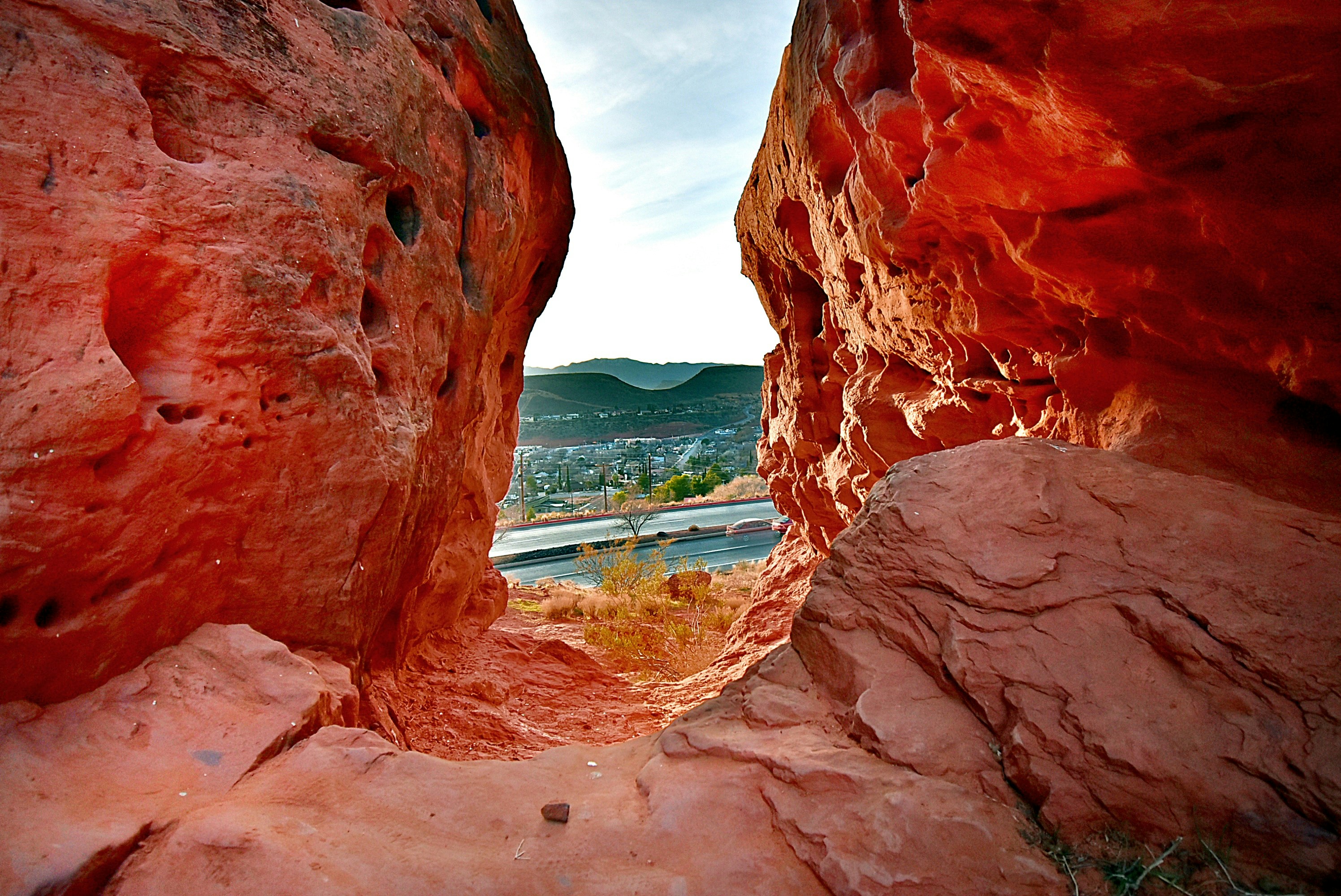 A natural rock formation frames a view of a valley below, showcasing vibrant red hues and distant hills under a clear sky.