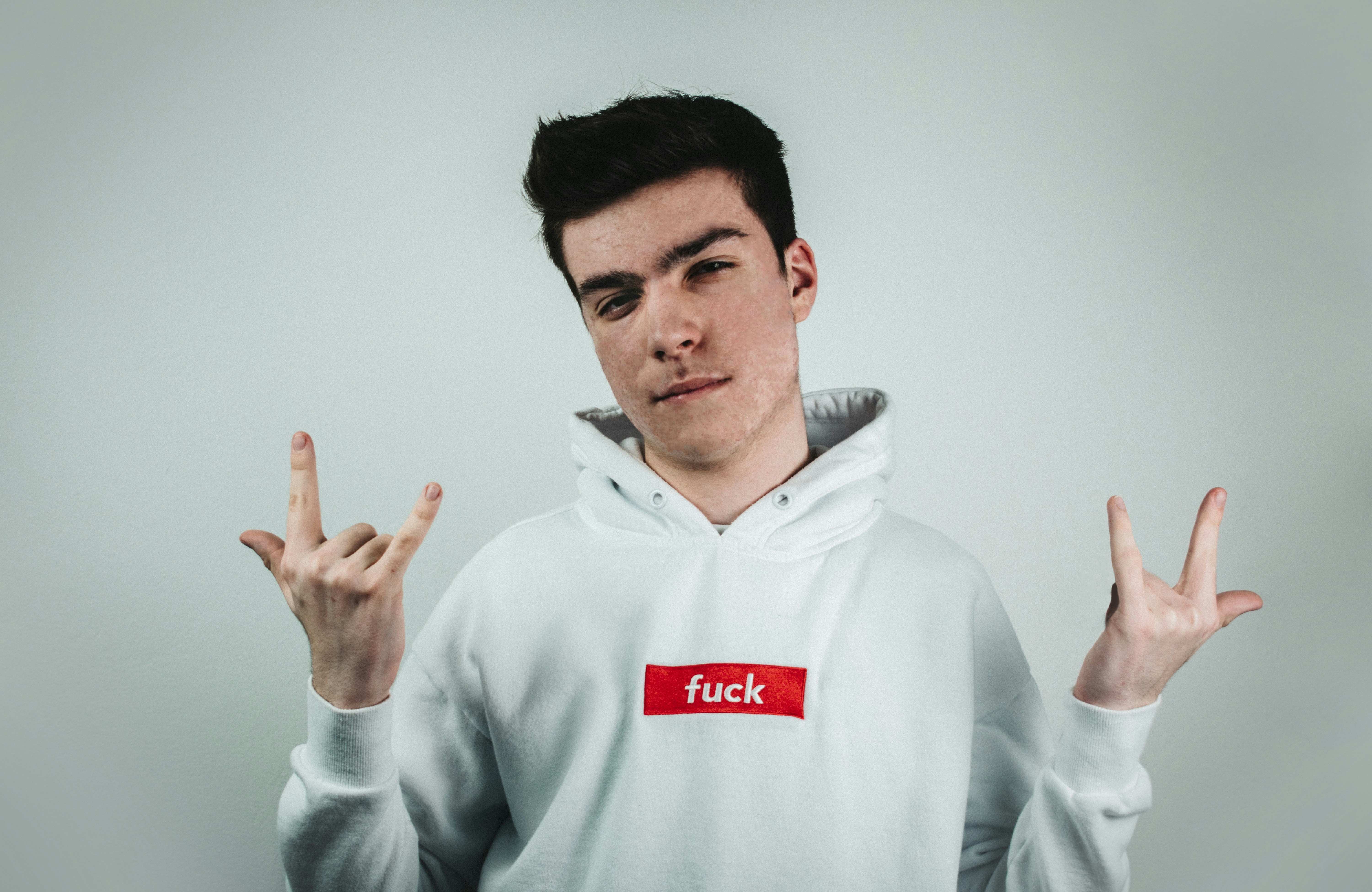 Young man in a white hoodie with a bold red logo strikes a rebellious pose against a neutral background.