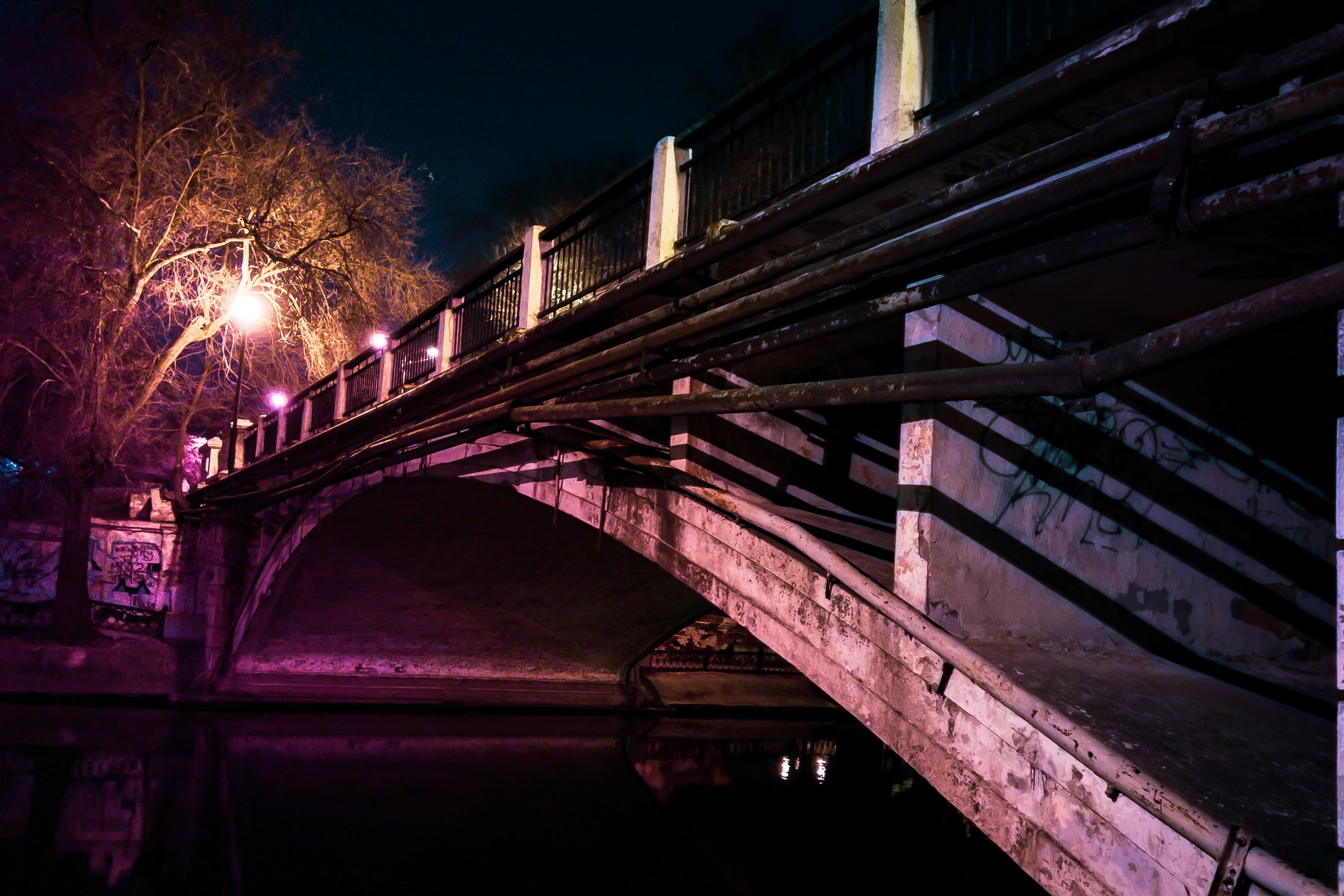 Night scene of a stone bridge spanning a calm river, bathed in pink-purple lights. The diagonal railing and its reflection create a moody leading line.