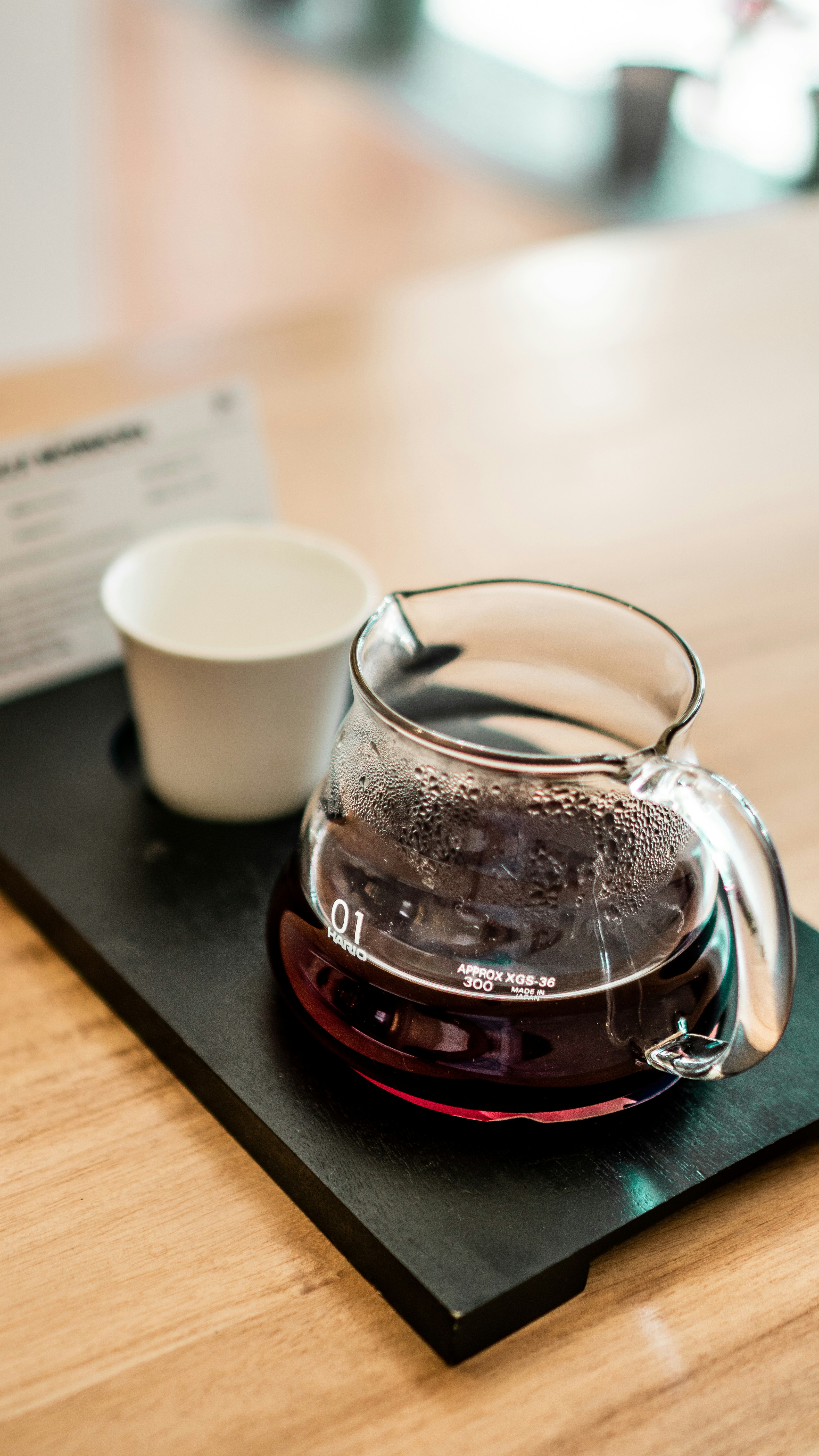 Glass coffee carafe filled with dark liquid sits on a wooden table next to a small white cup. Steam rises from the carafe, hinting at the warmth of the brew.