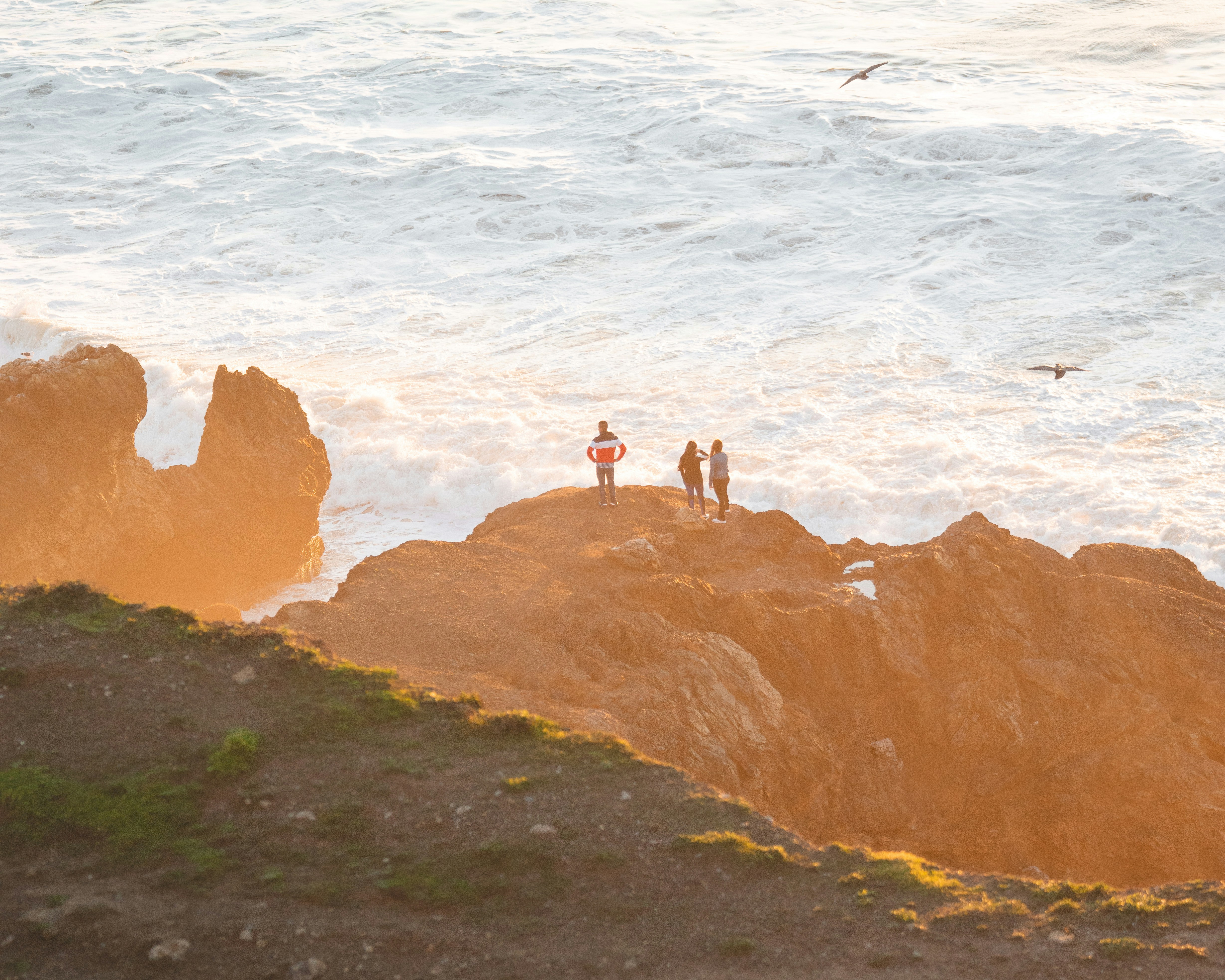 Three figures standing on a rocky outcrop, gazing at the crashing waves below, with sunlight casting a warm glow over the scene.