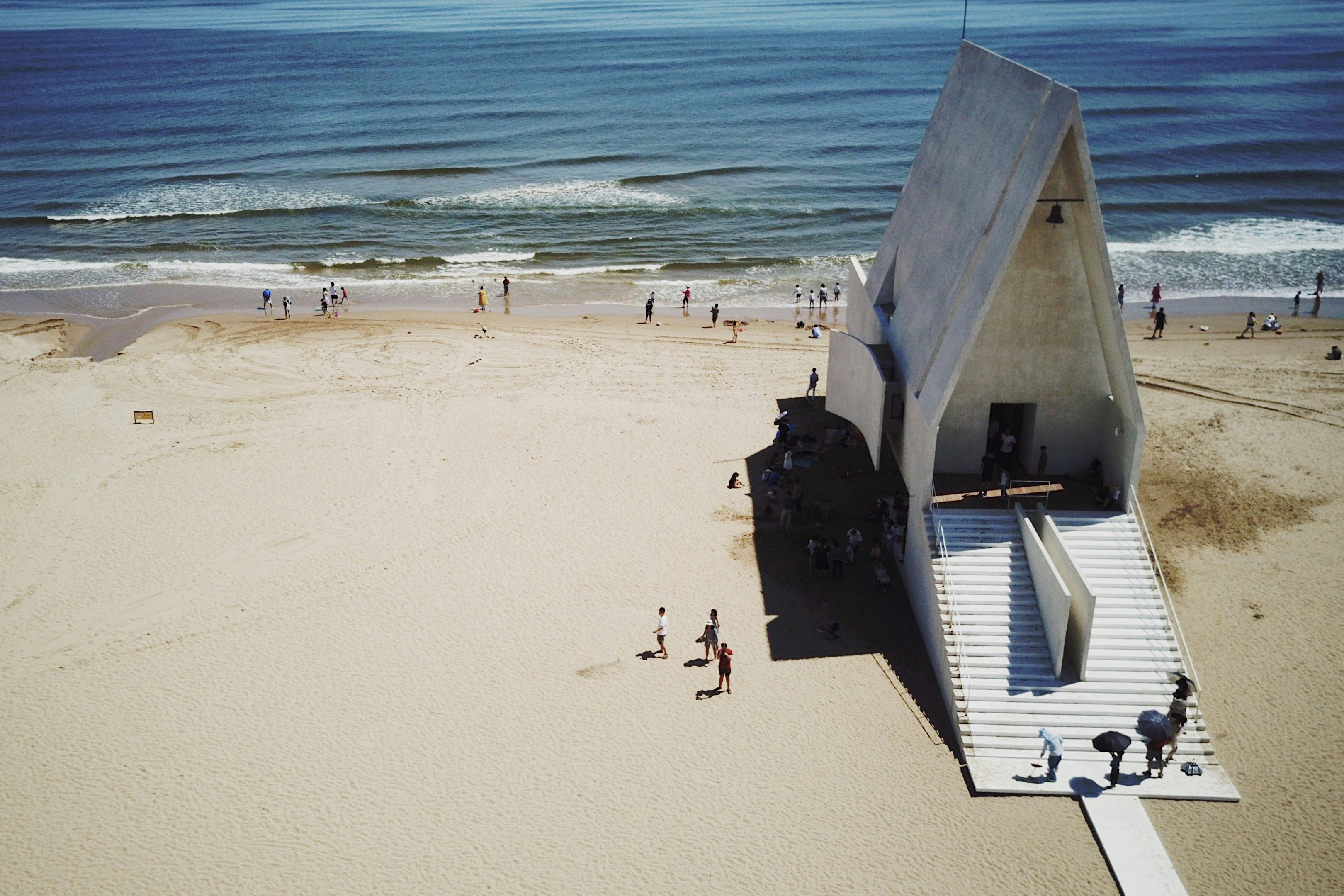 Unique modern chapel set against a serene beach backdrop with people enjoying the sandy shore.