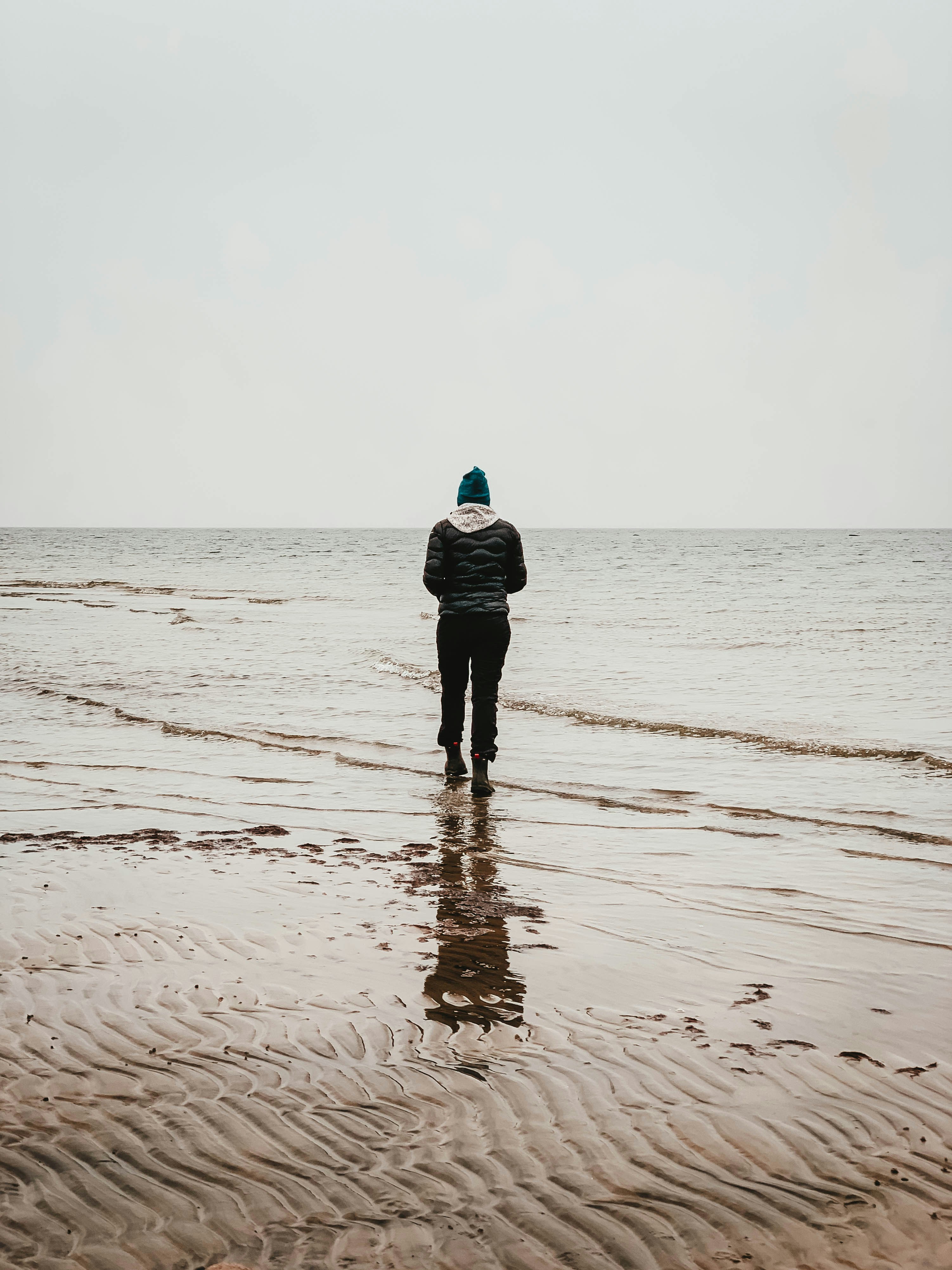 Person walking along a wet sandy beach, reflecting on the calm sea under a gray sky.