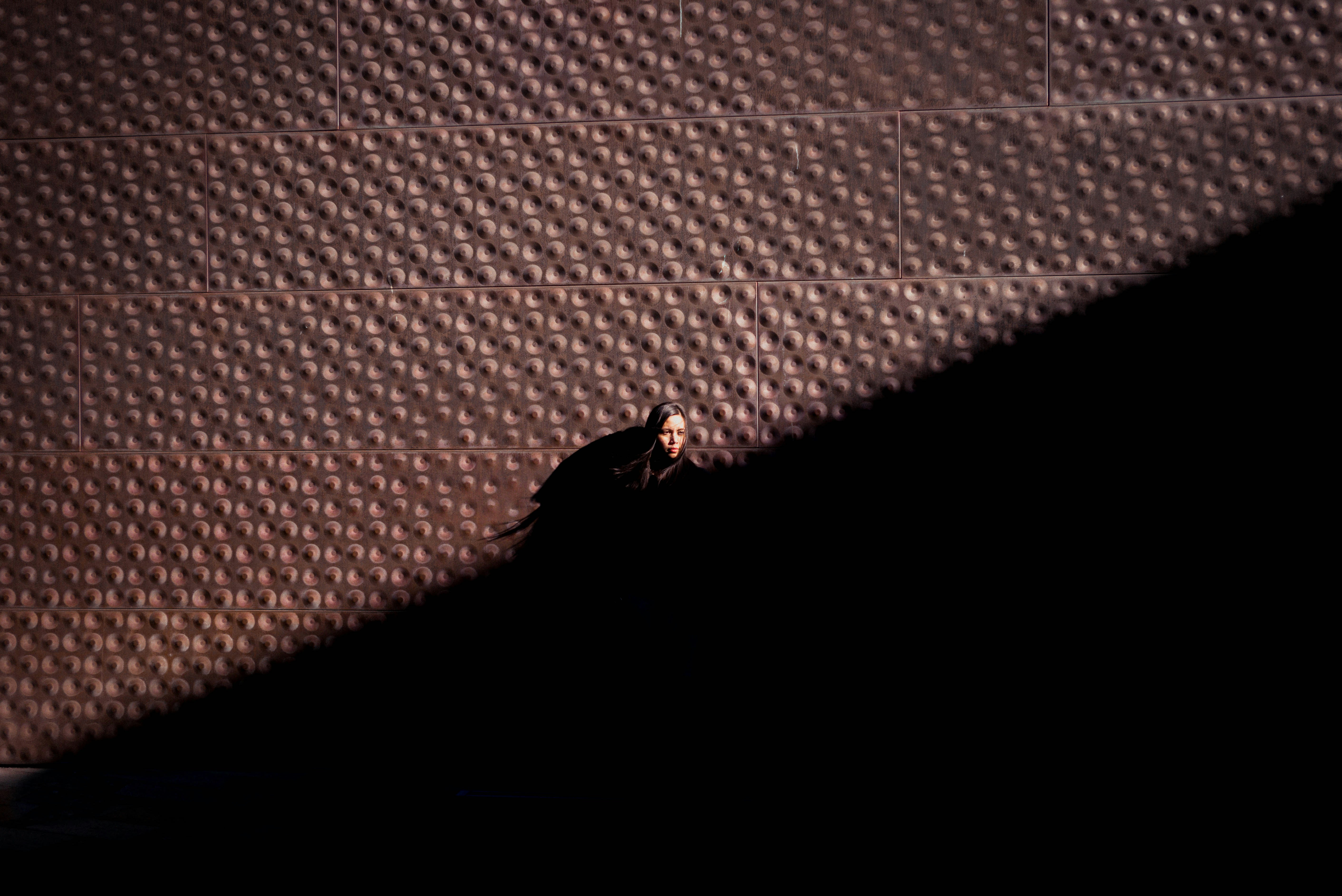 A figure stands against a textured wall, partially obscured by a dramatic shadow, highlighting the interplay of light and form.