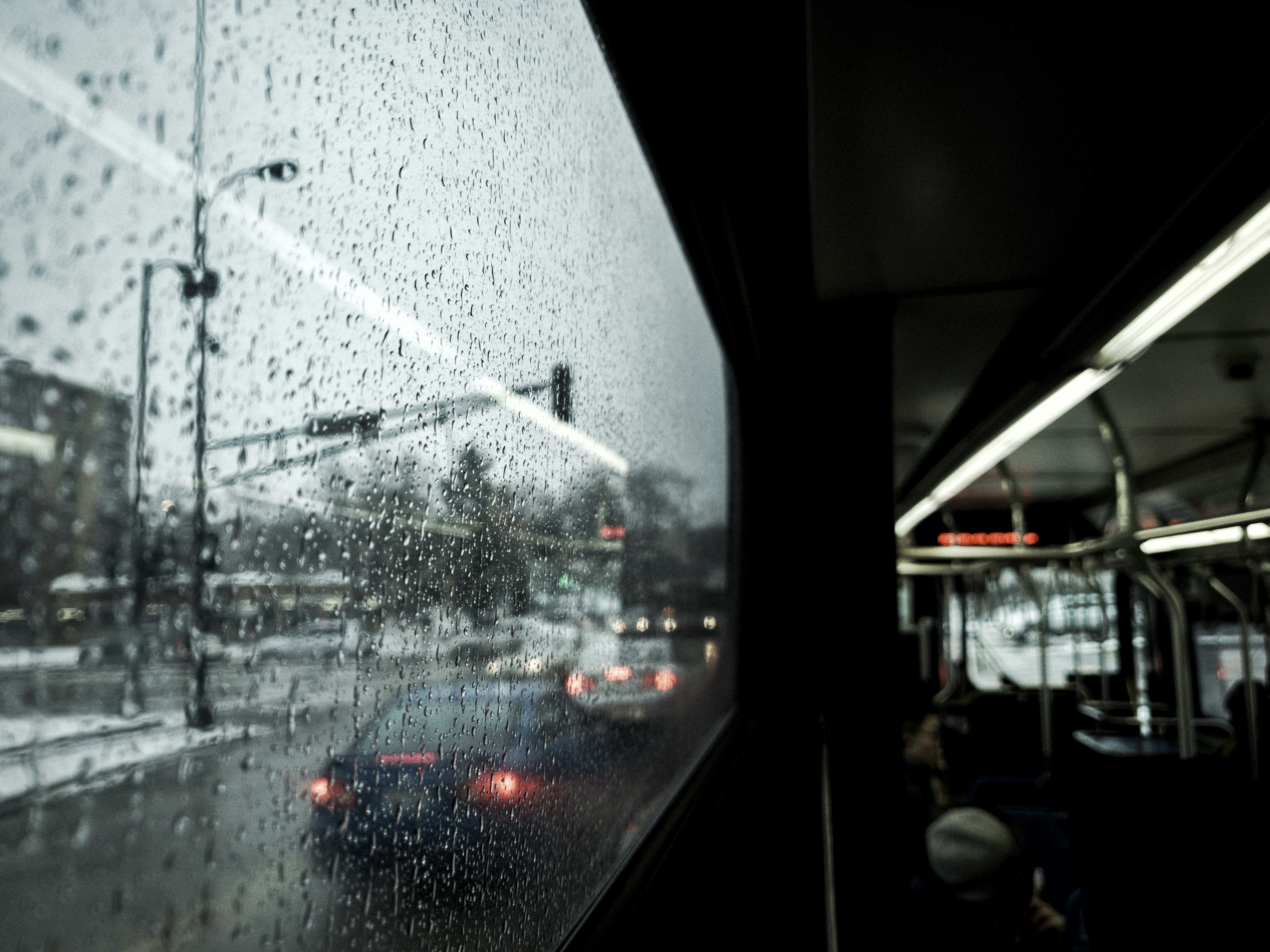 Raindrops on a bus window blur the view of a city street with cars and traffic lights under a gray sky.