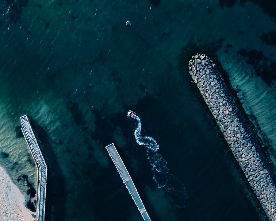 Aerial view of a small boat cutting through dark water between stone breakwaters and dock structures. White wake curves behind it near a sandy shoreline.