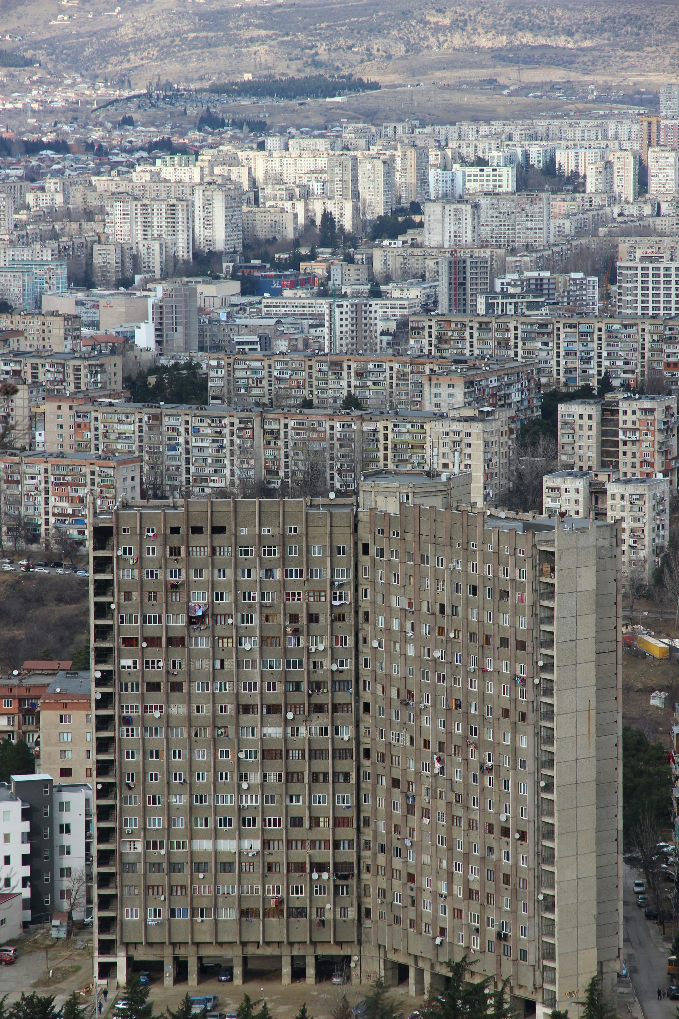 A high-rise apartment building stands prominently in the foreground, surrounded by a sprawling cityscape of uniform structures and distant mountains.