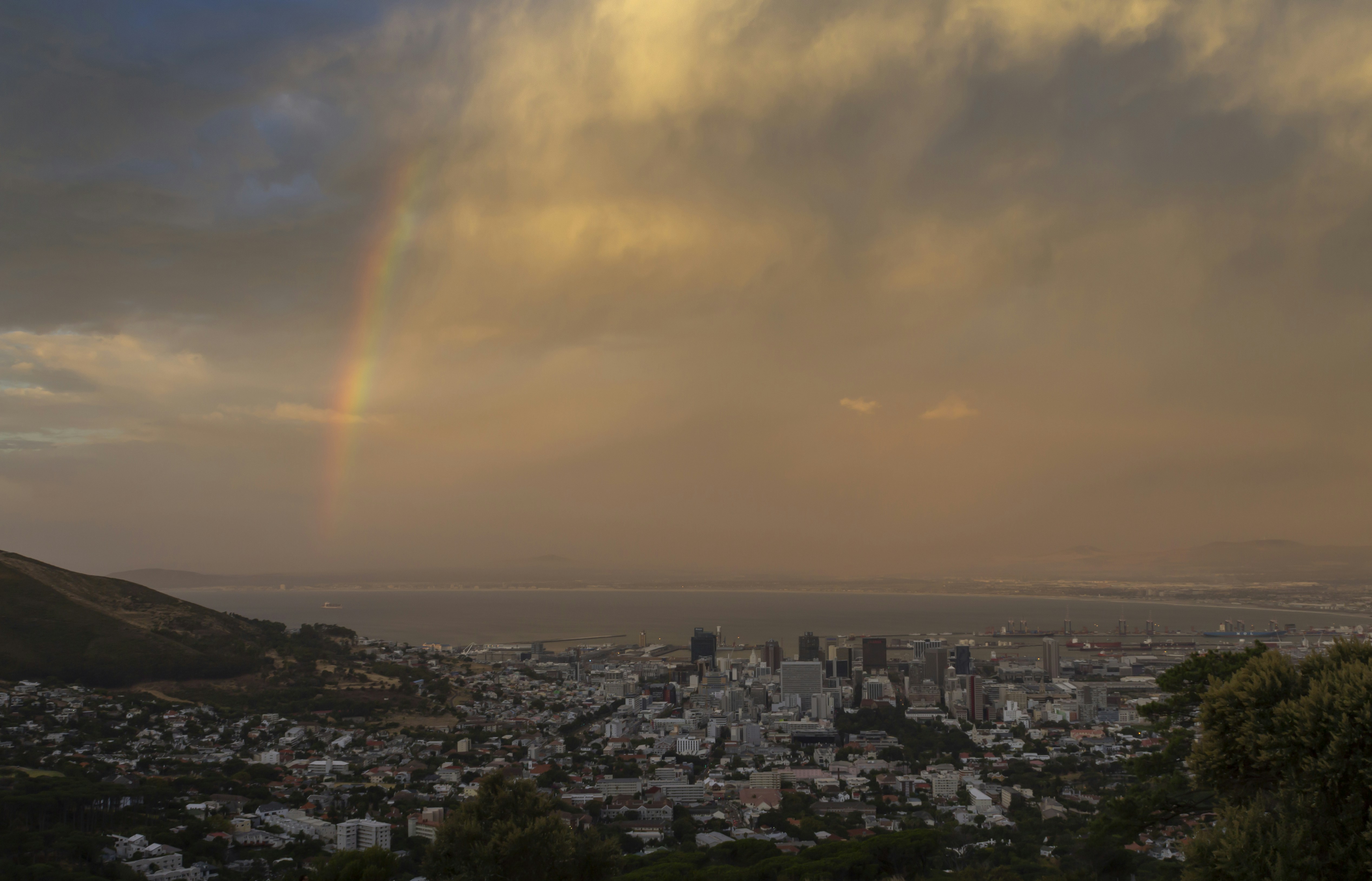 A vibrant rainbow arches over a sprawling city, framed by dramatic clouds and the distant ocean. The scene captures the interplay of light and urban life.