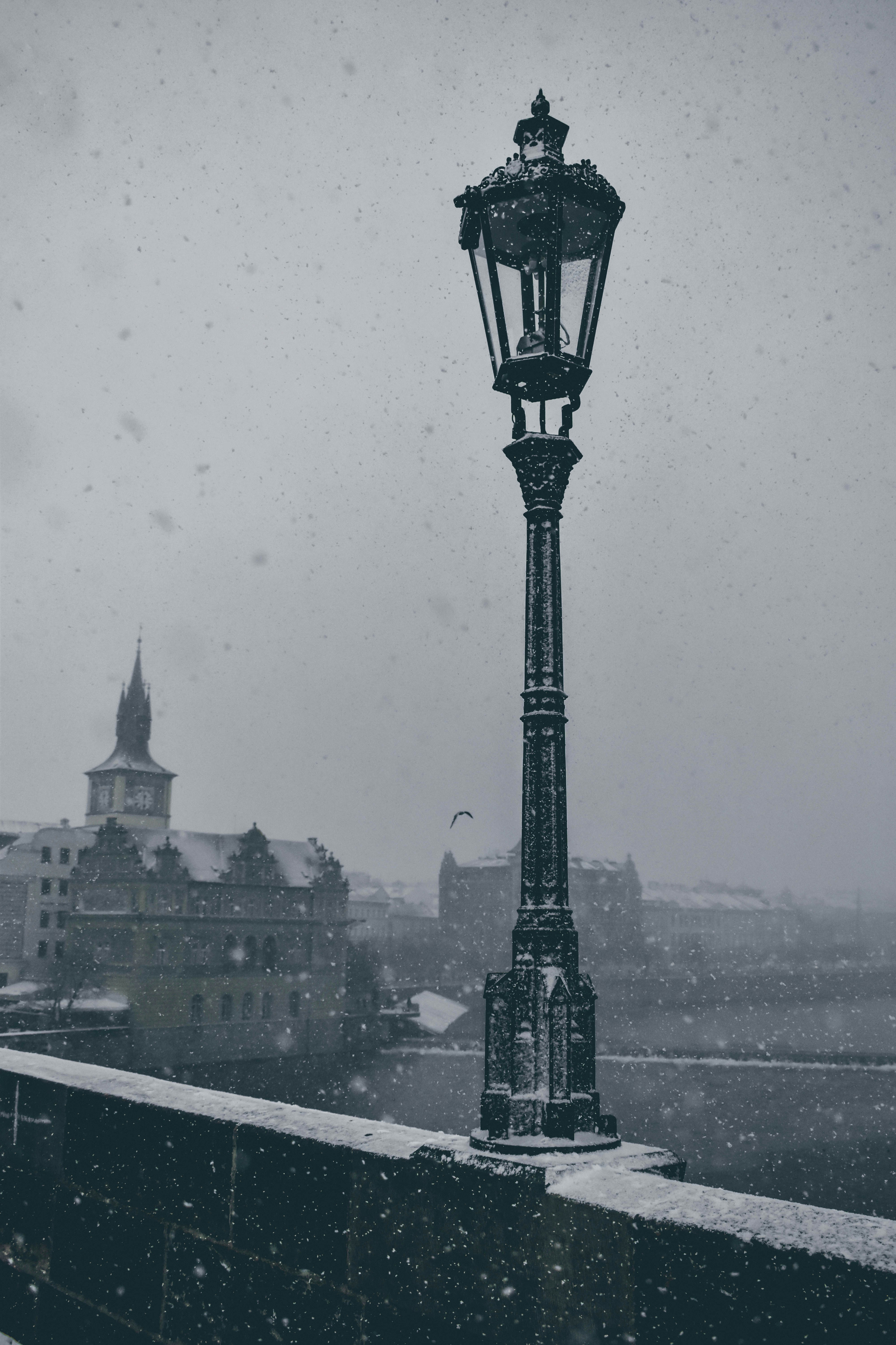 A vintage street lamp stands resolute against a snowy backdrop, highlighting the architecture of distant buildings as snowflakes dance in the air.