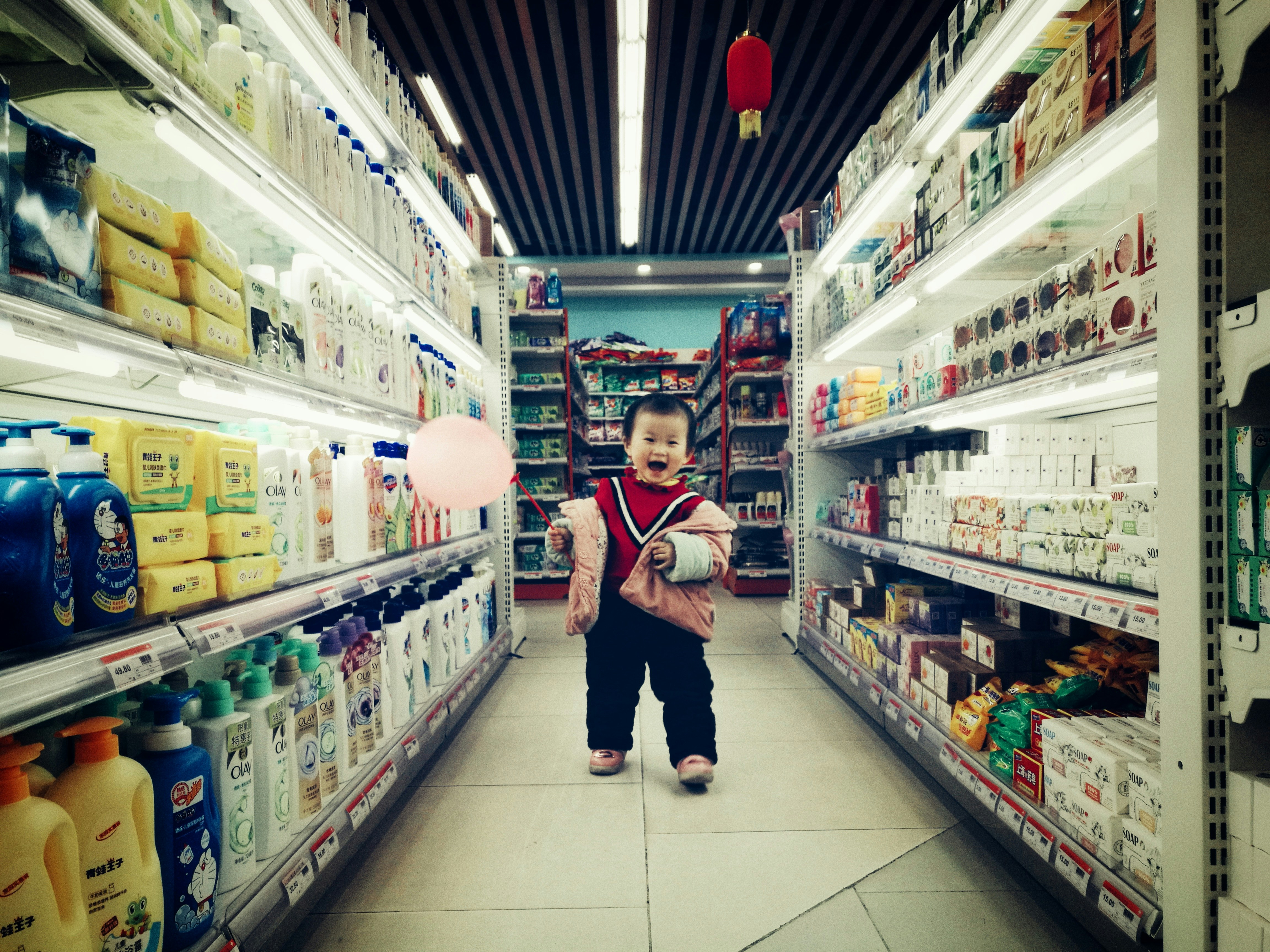 Child holding a pink balloon while standing between shelves filled with various products in a grocery store.