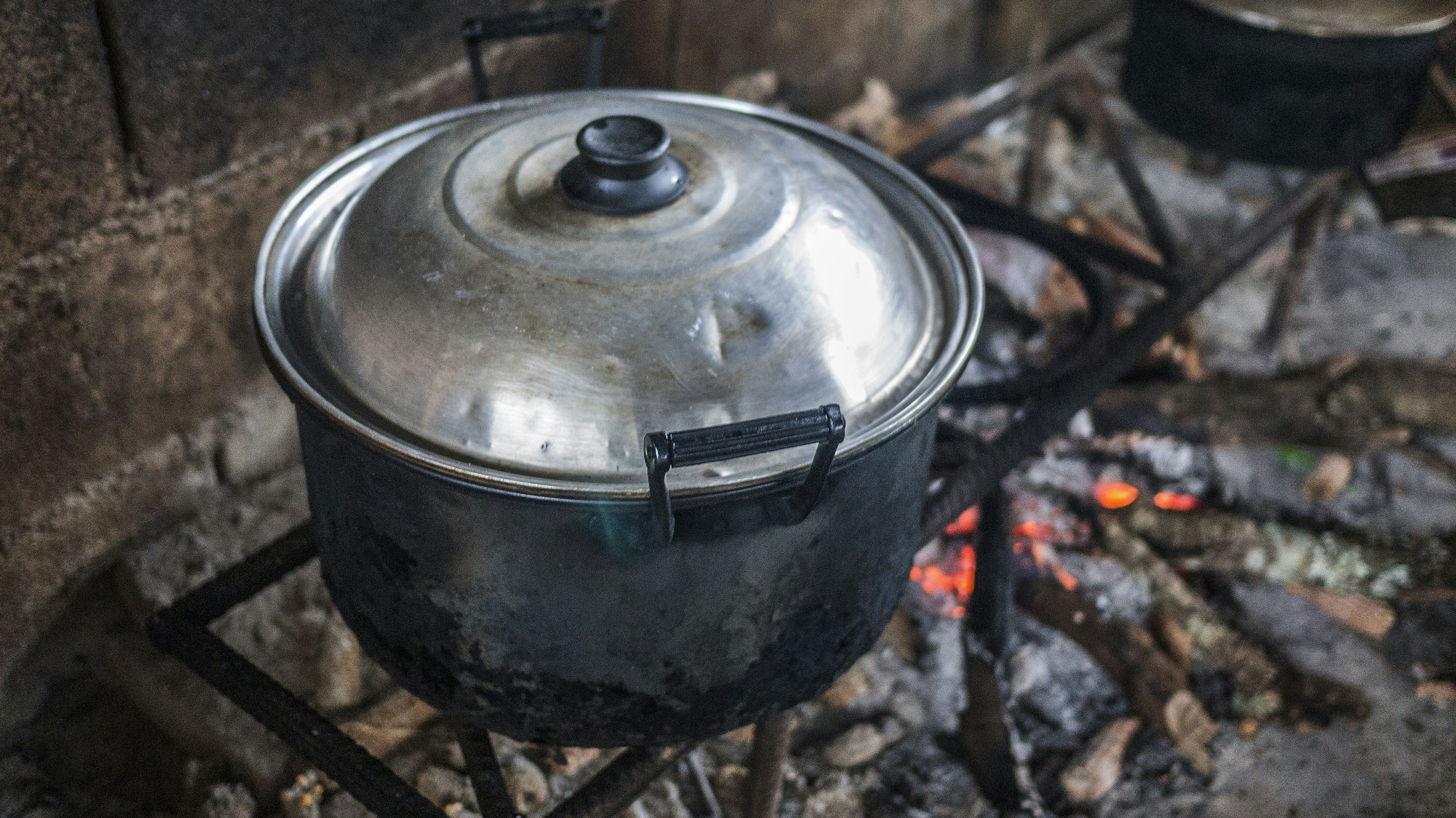 A large metal pot simmering over an open flame on a rustic stove, surrounded by ashes and embers. The scene evokes a sense of traditional cooking methods.