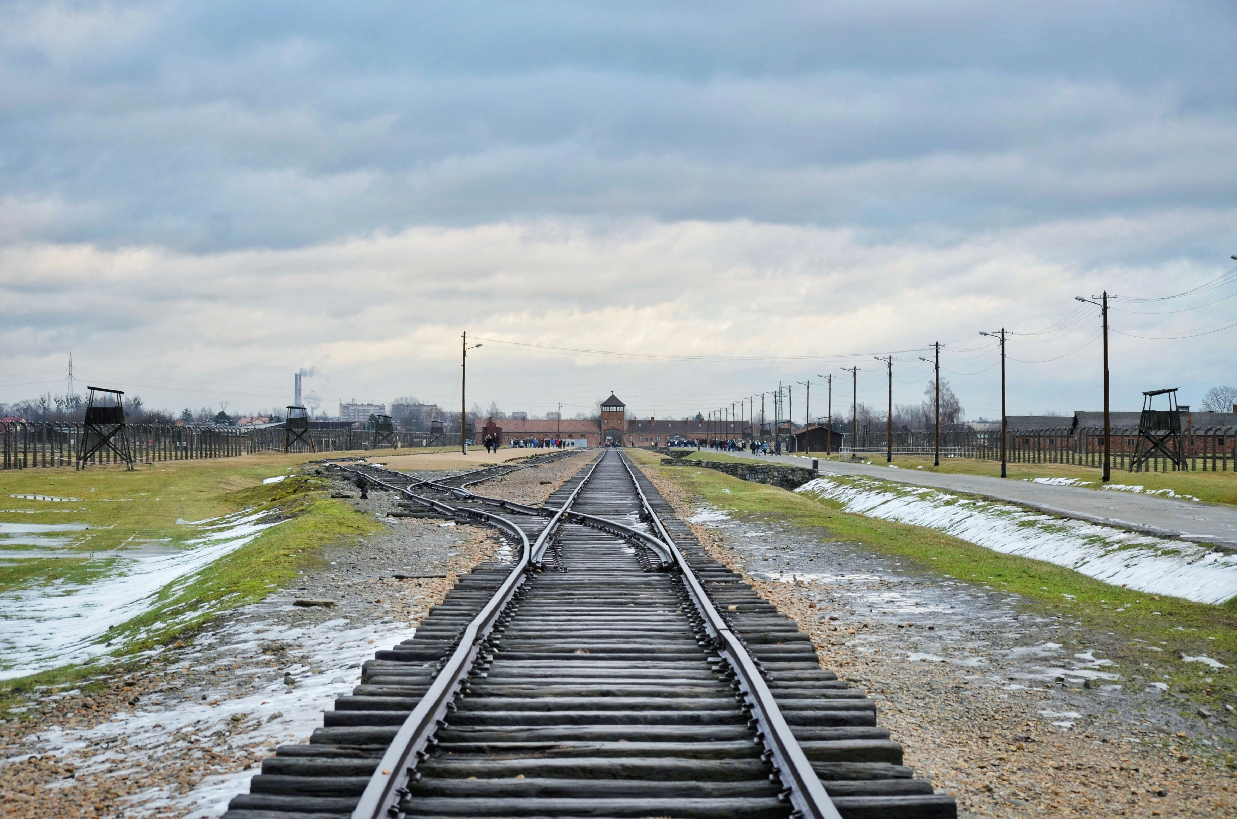 Railway tracks stretching towards a distant horizon under a cloudy sky, bordered by patches of snow and grass.