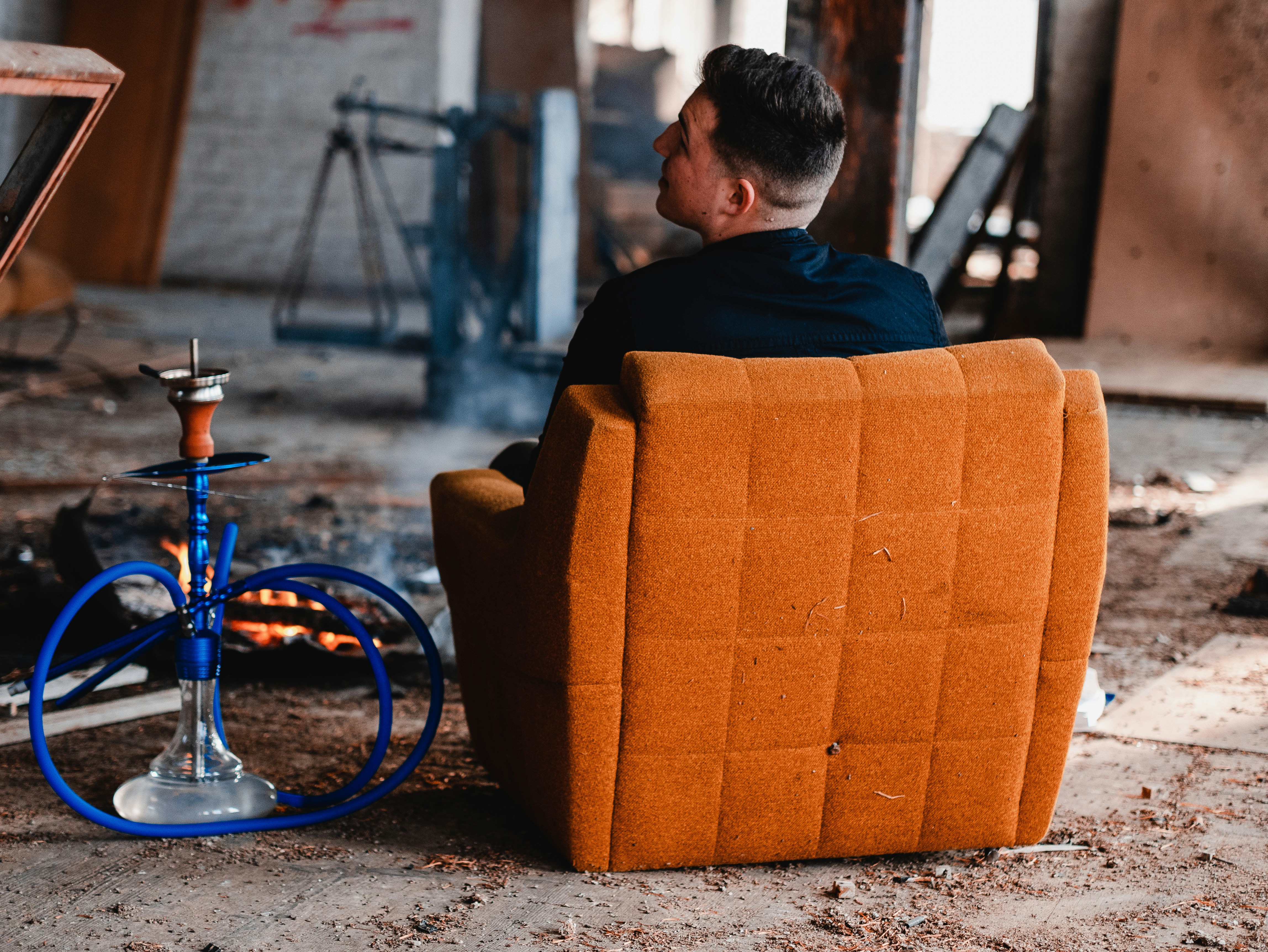 A man sits in an orange armchair, gazing thoughtfully at a hookah beside a smoldering fire in an abandoned building.