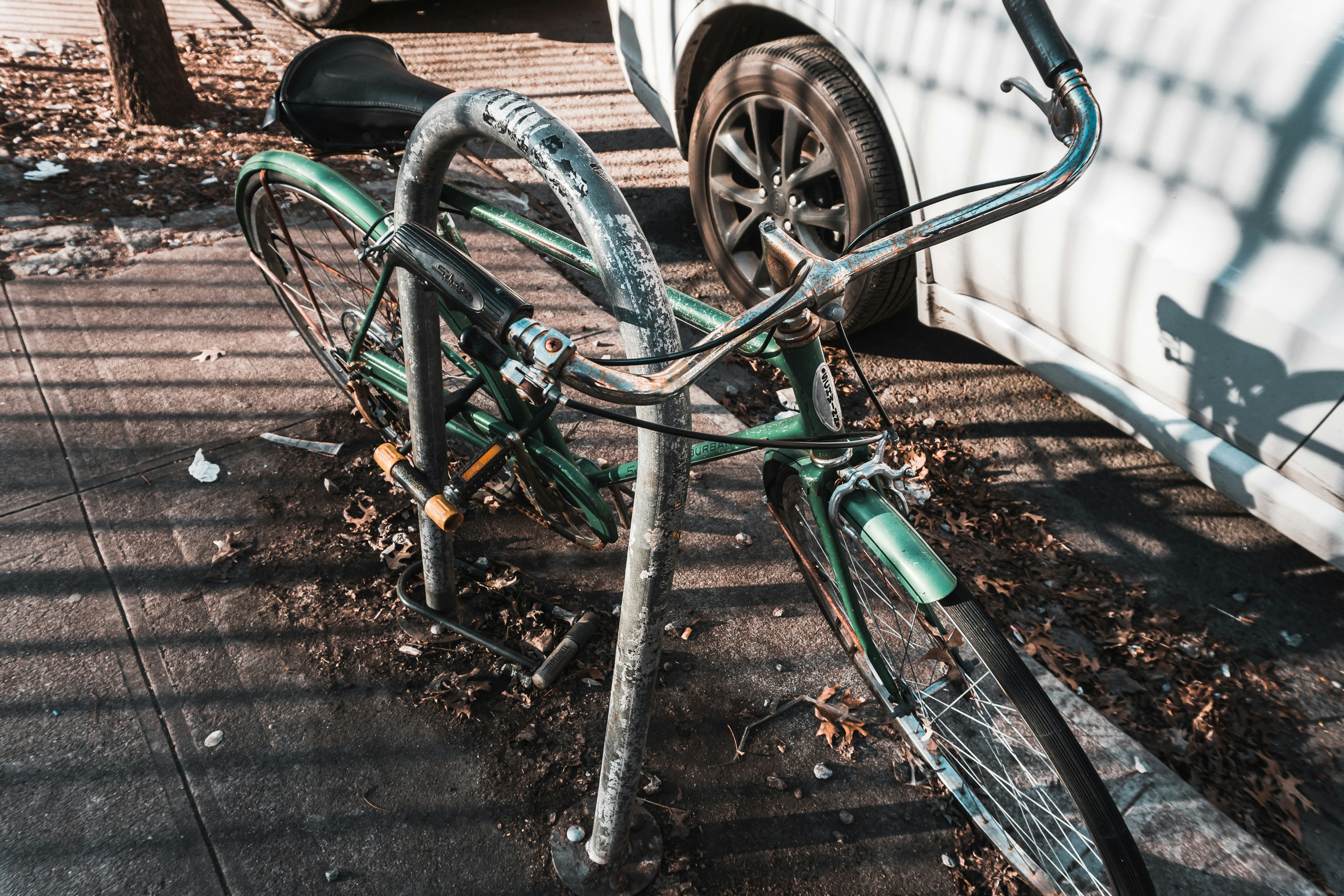 A vintage bicycle locked to a weathered rack, surrounded by fallen leaves and urban debris. The scene reflects a blend of neglect and character in the city.