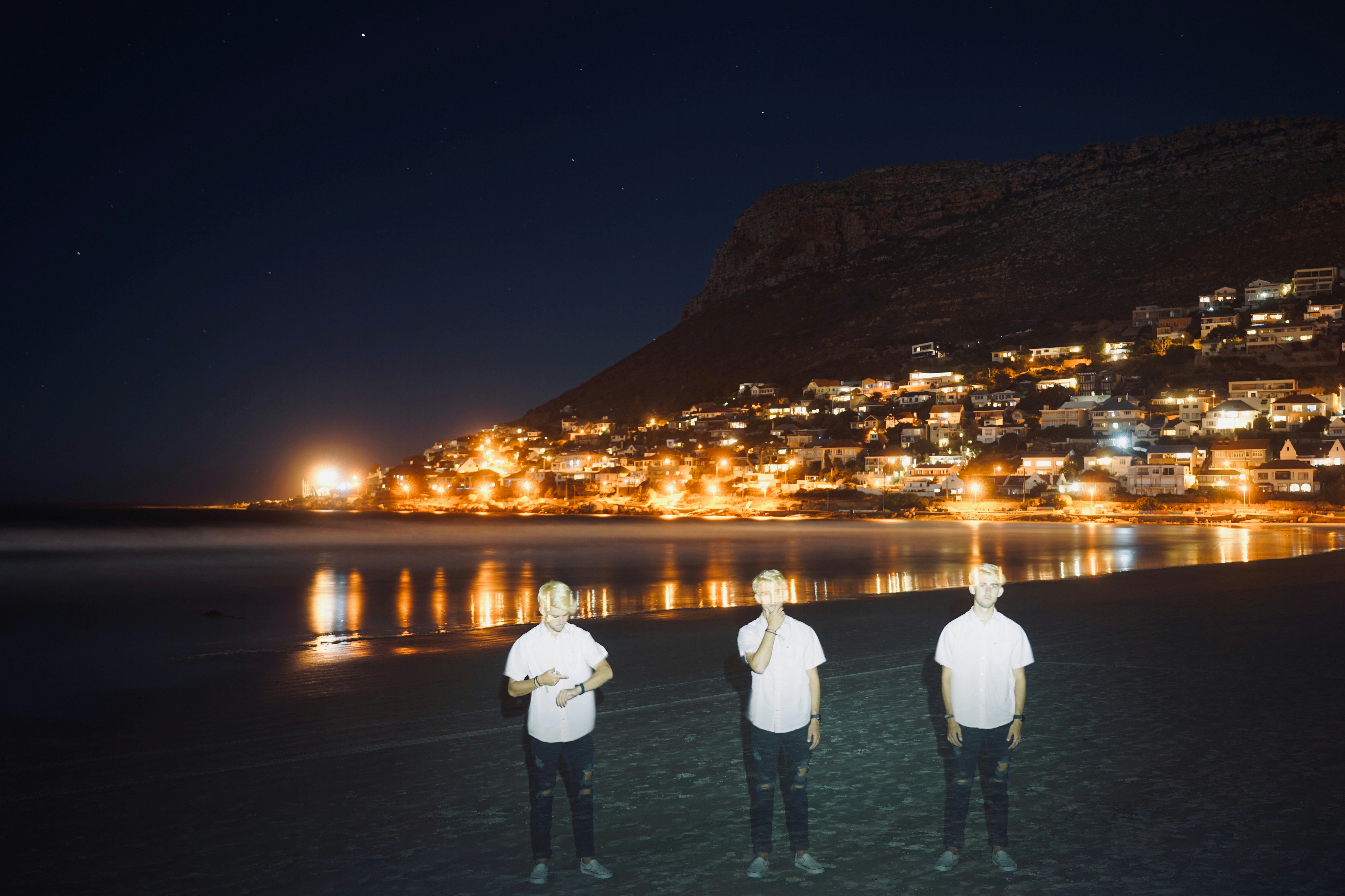 Three figures stand on a beach at night, illuminated by distant city lights and stars above. The calm water reflects the vibrant glow of the coastal town.