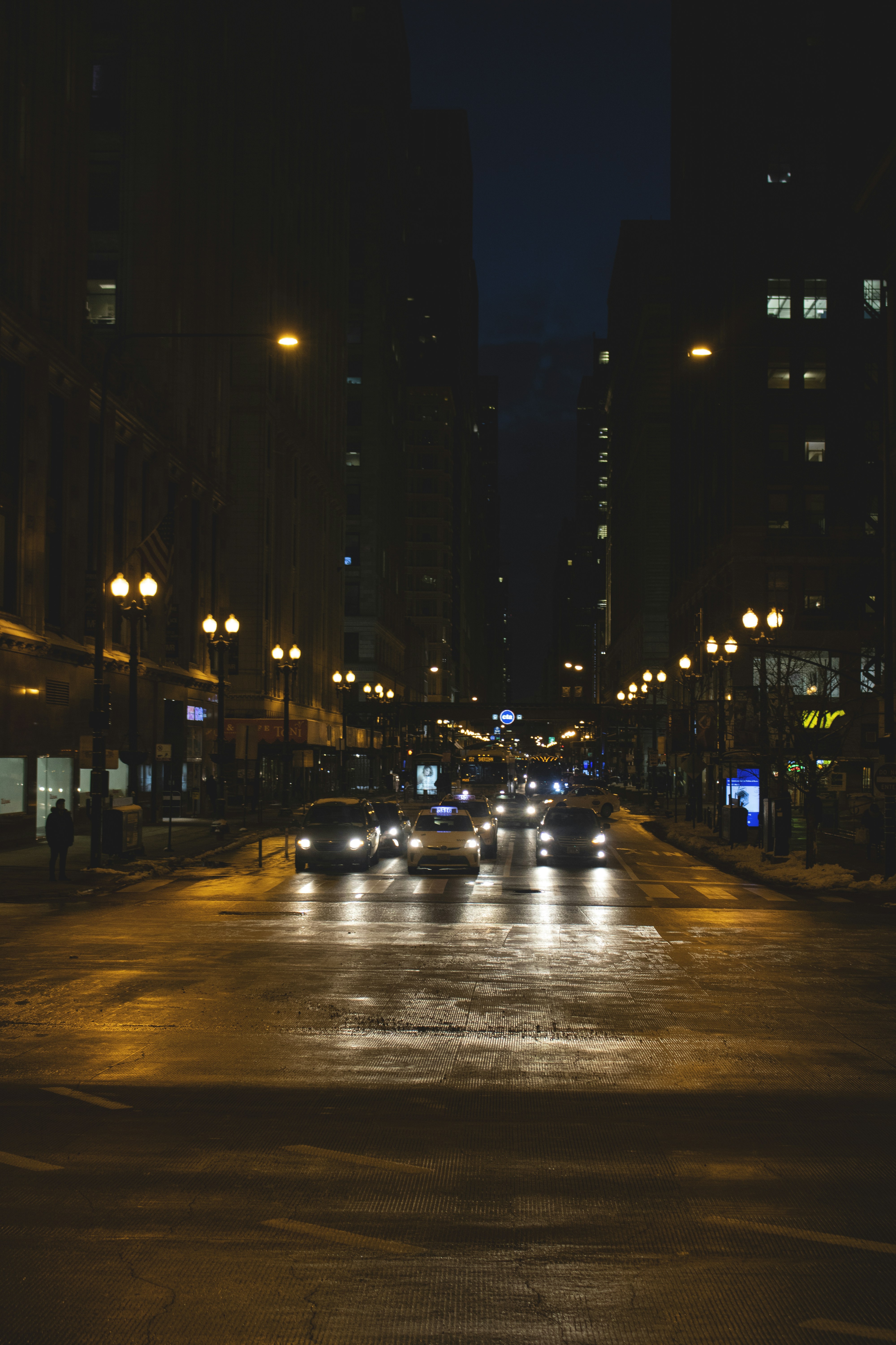 Illuminated street scene at night with cars navigating a bustling urban environment, featuring glowing streetlights and reflections on wet pavement.