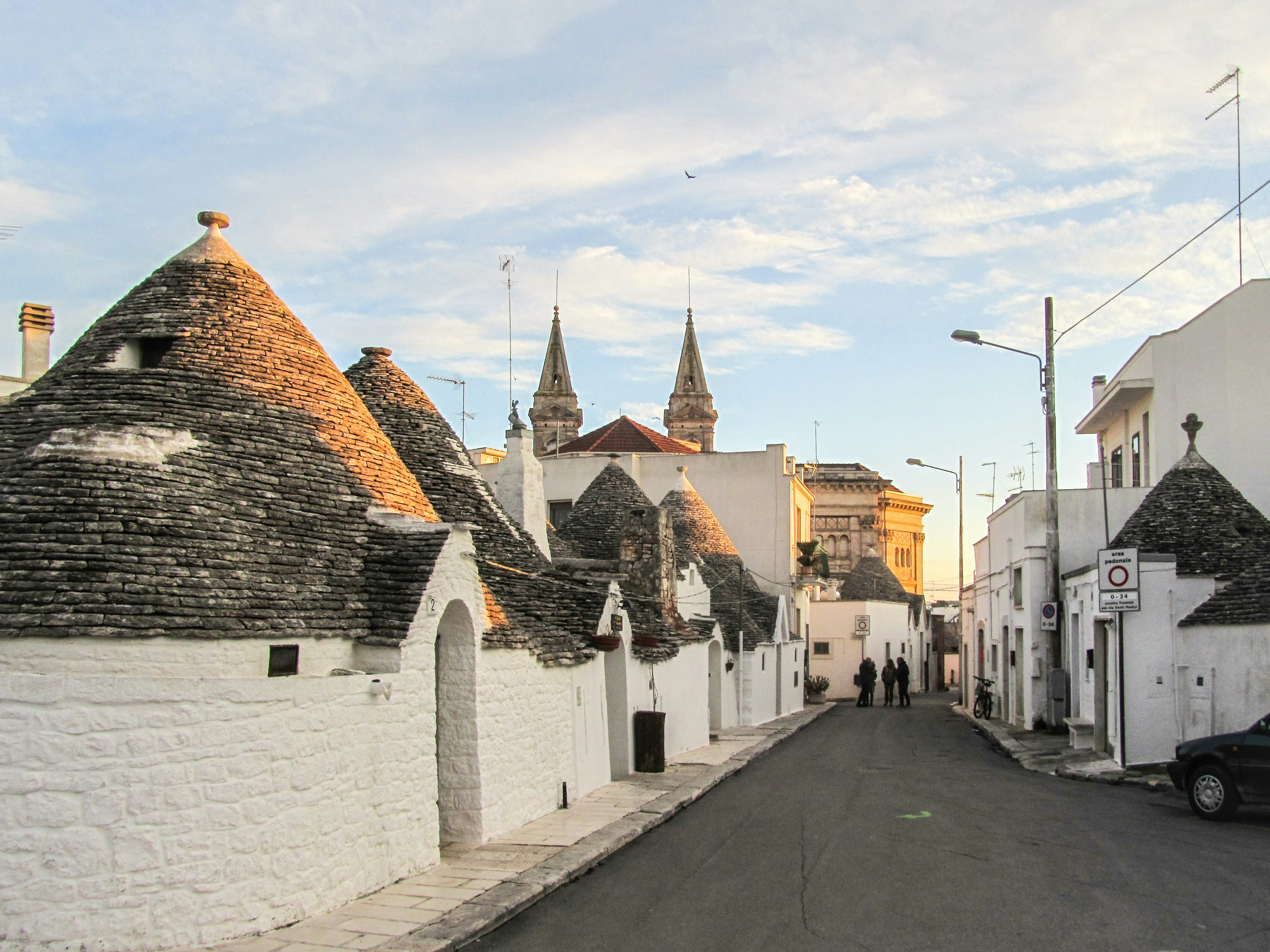 Traditional trulli houses with conical roofs line a narrow street under a partly cloudy sky.