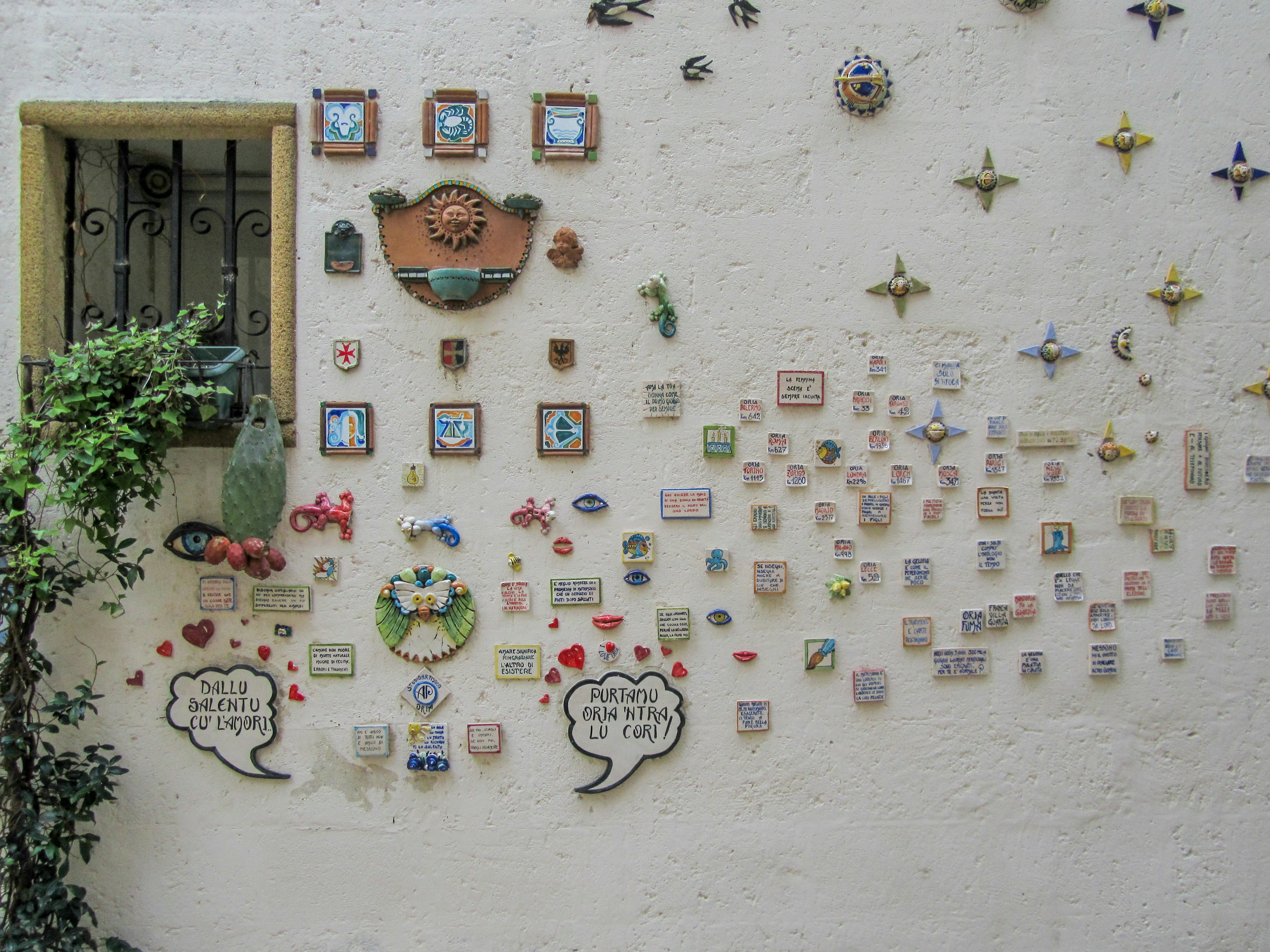 Artistic arrangement of colorful ceramic tiles and decorations on a white wall beside a small window adorned with greenery.