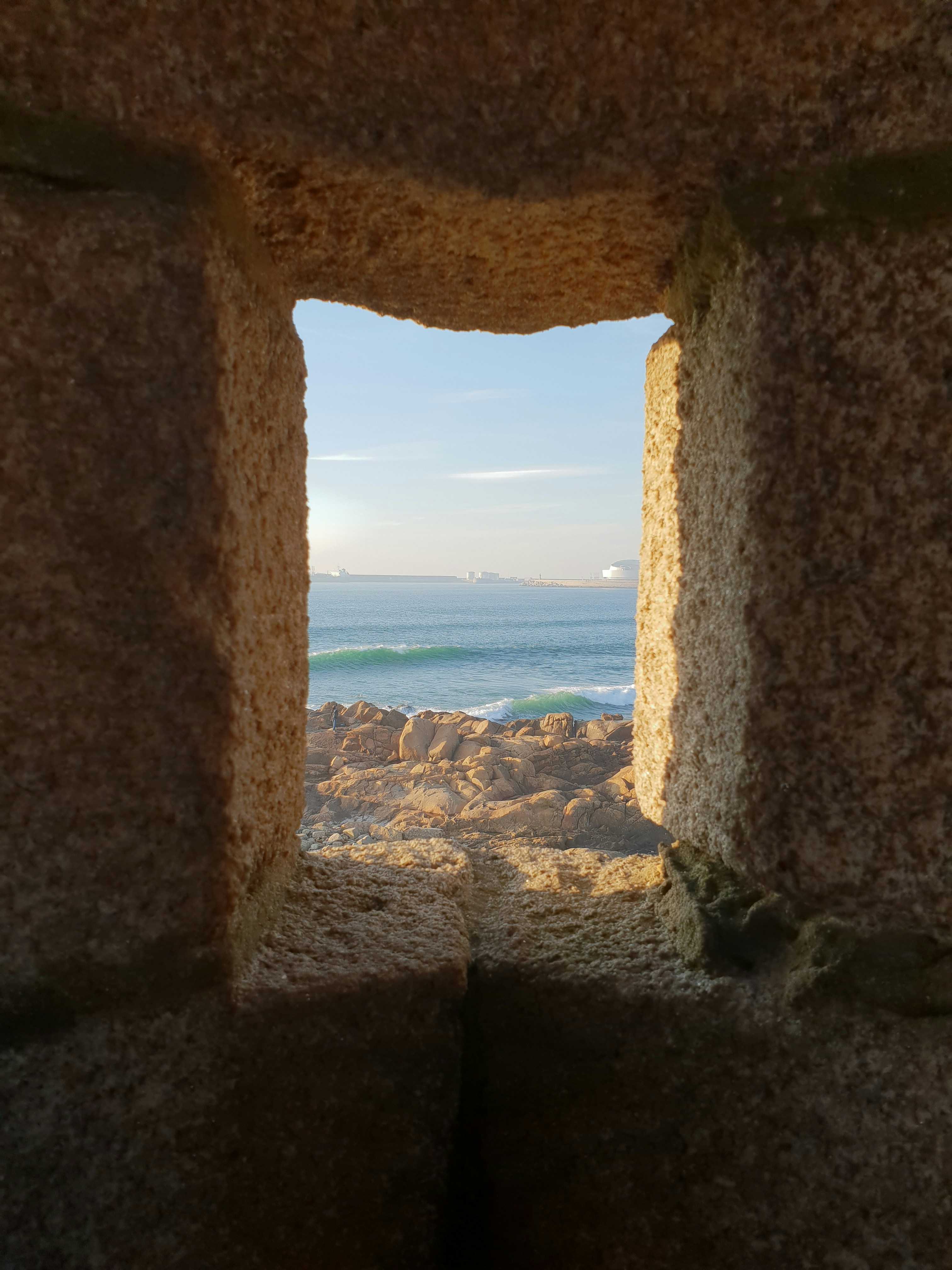 View of a tranquil ocean scene through a stone window, highlighting the contrast between rugged rock and gentle waves. The soft light enhances the serene atmosphere.
