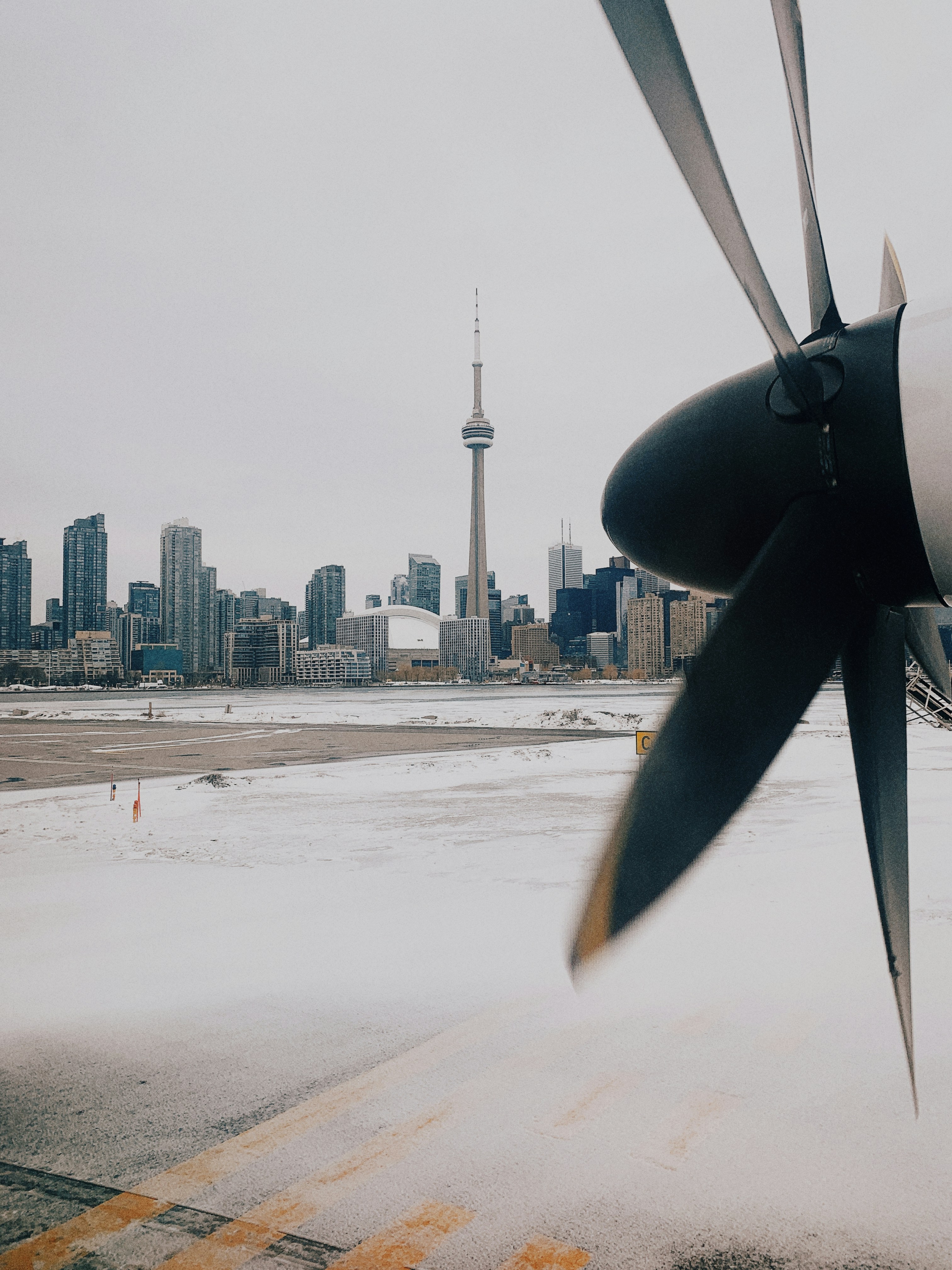 Propeller of an aircraft in the foreground with Toronto's CN Tower and skyline visible in the background, set against a cloudy sky.