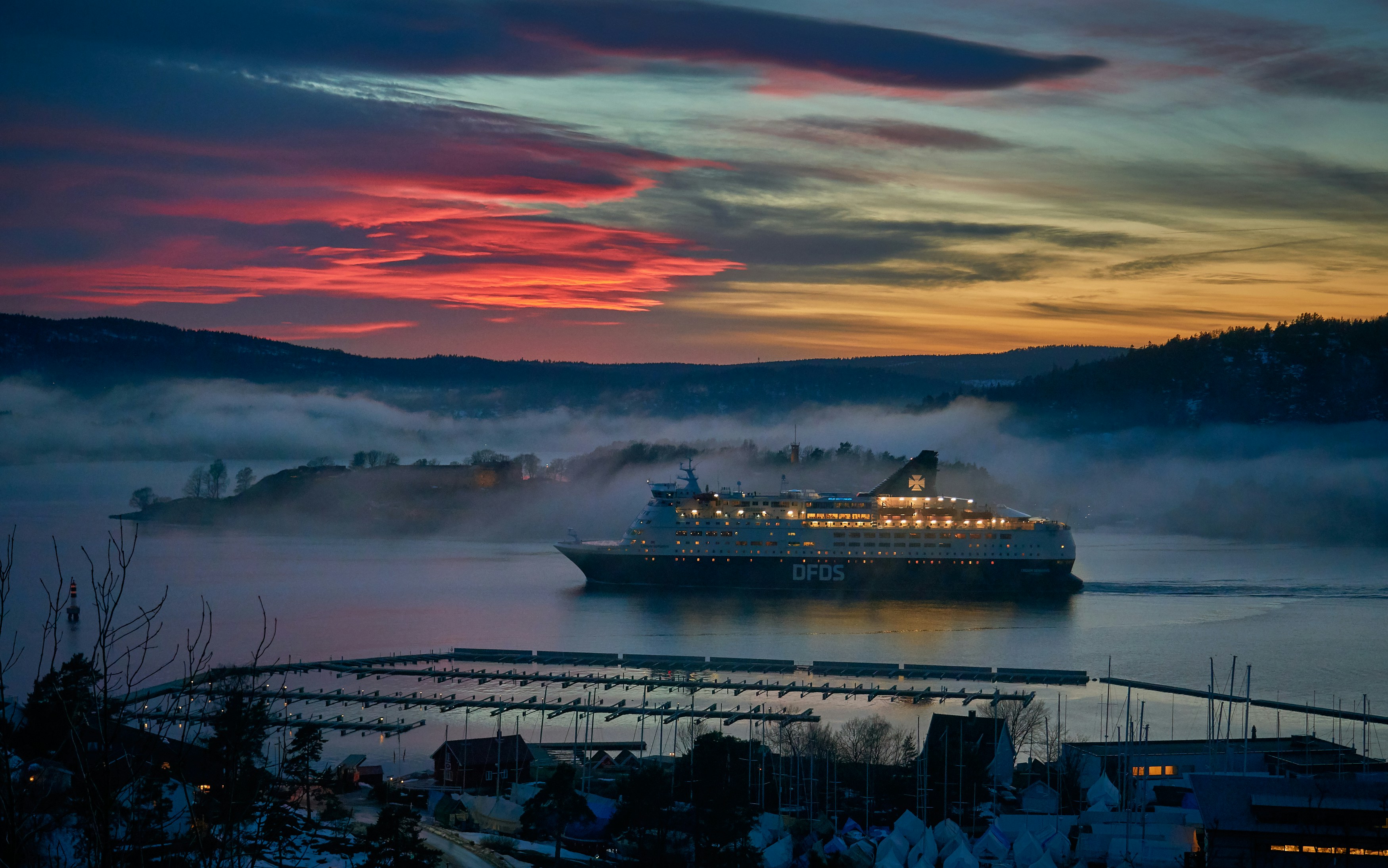 A cruise ship glides through foggy waters at dusk, illuminated by warm lights against a backdrop of vibrant sunset colors.
