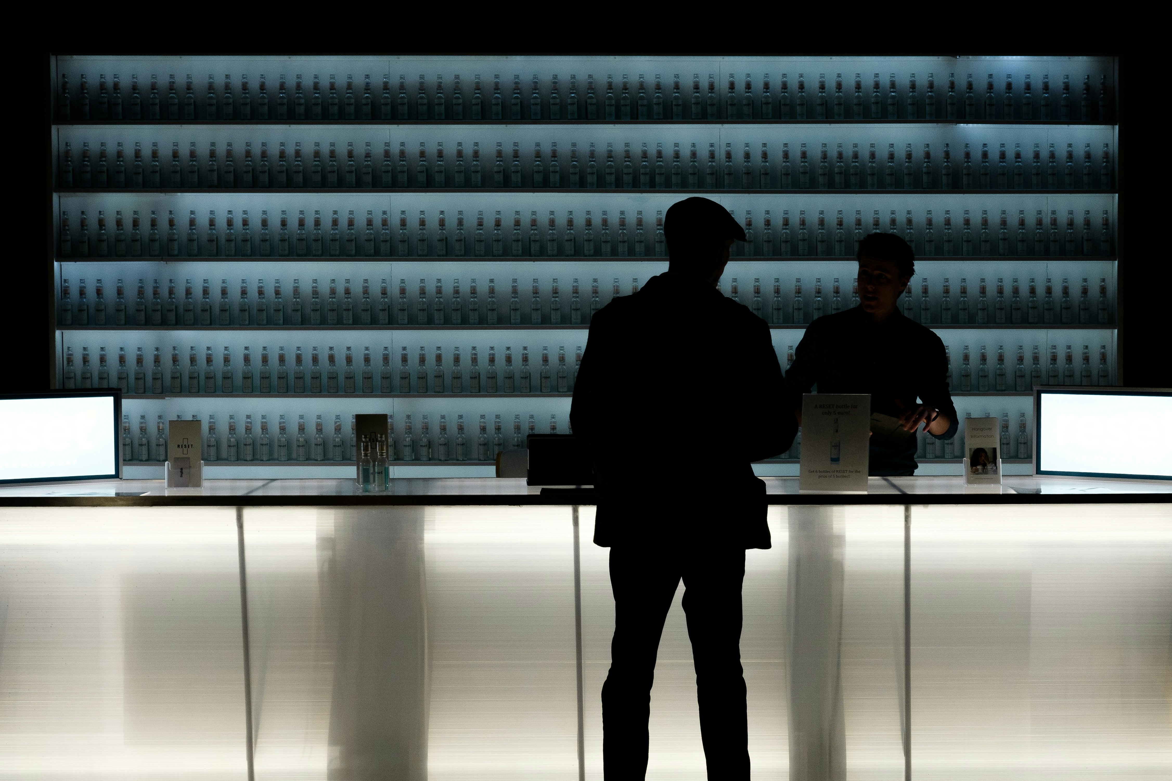 Two silhouettes converse at a sleek counter illuminated by soft backlighting, showcasing a modern bar atmosphere.