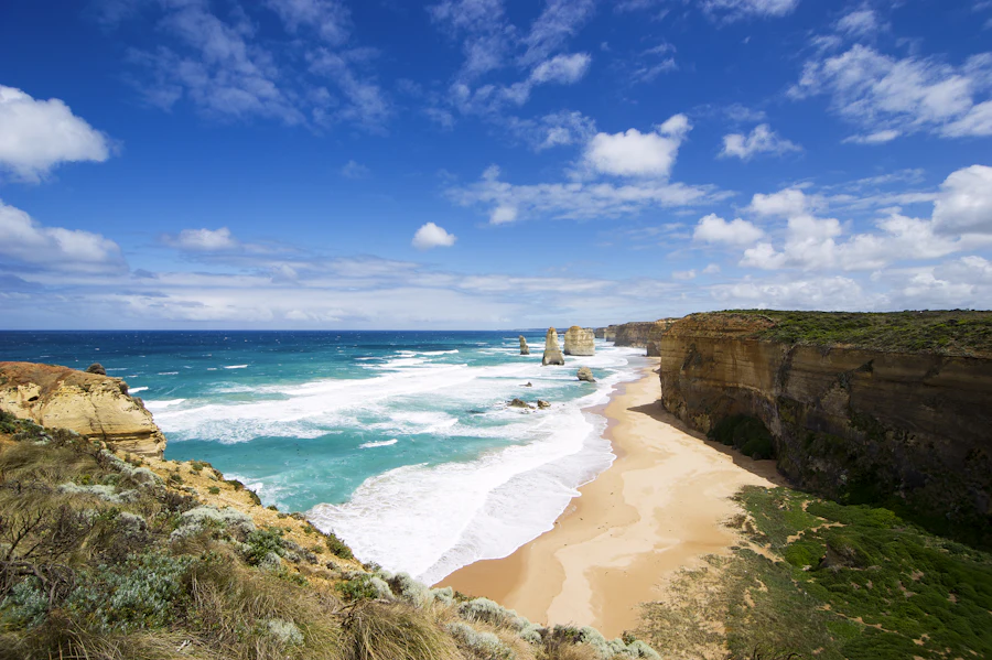 Bright coastal scene with turquoise waves rolling onto a sandy cove beneath tall limestone cliffs and a wide blue sky. The shoreline curves toward sea stacks in the distance.