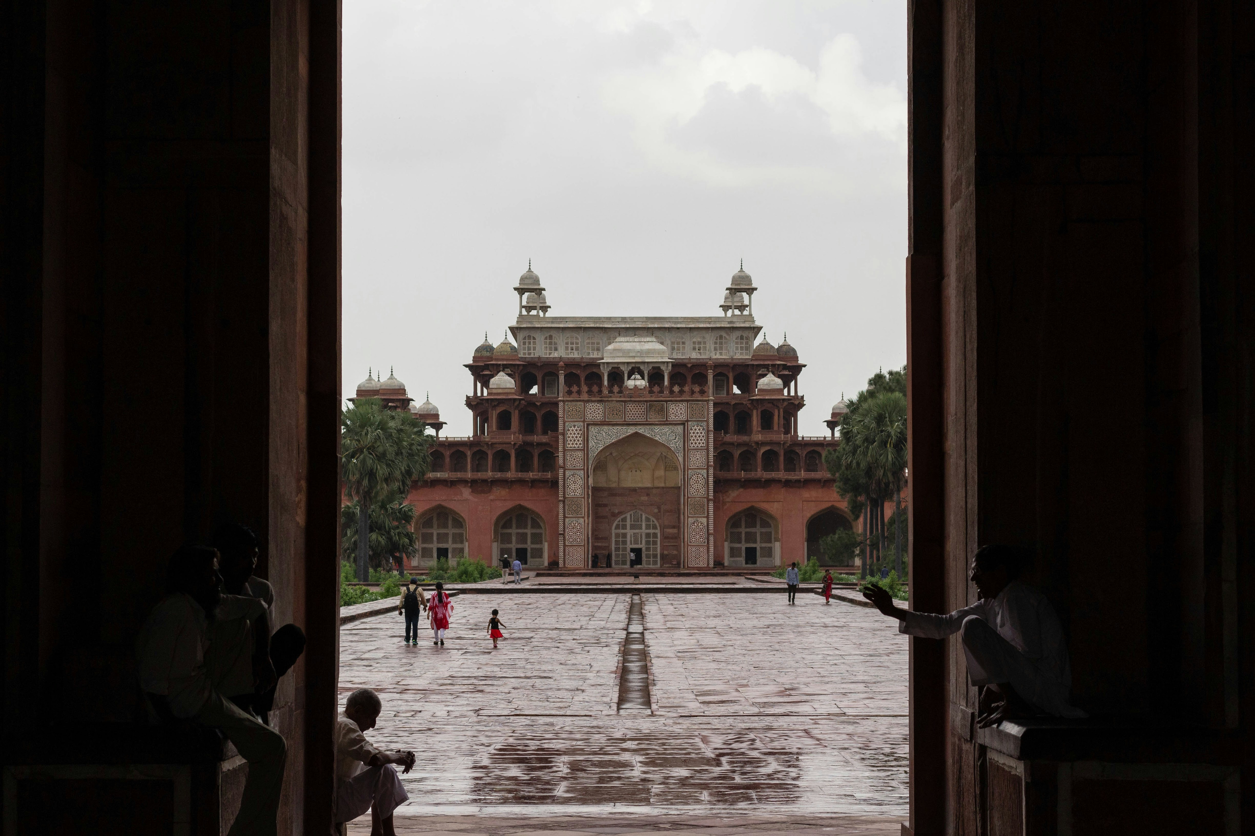 Archway framing a distant historic building with people walking in a courtyard on a cloudy day.