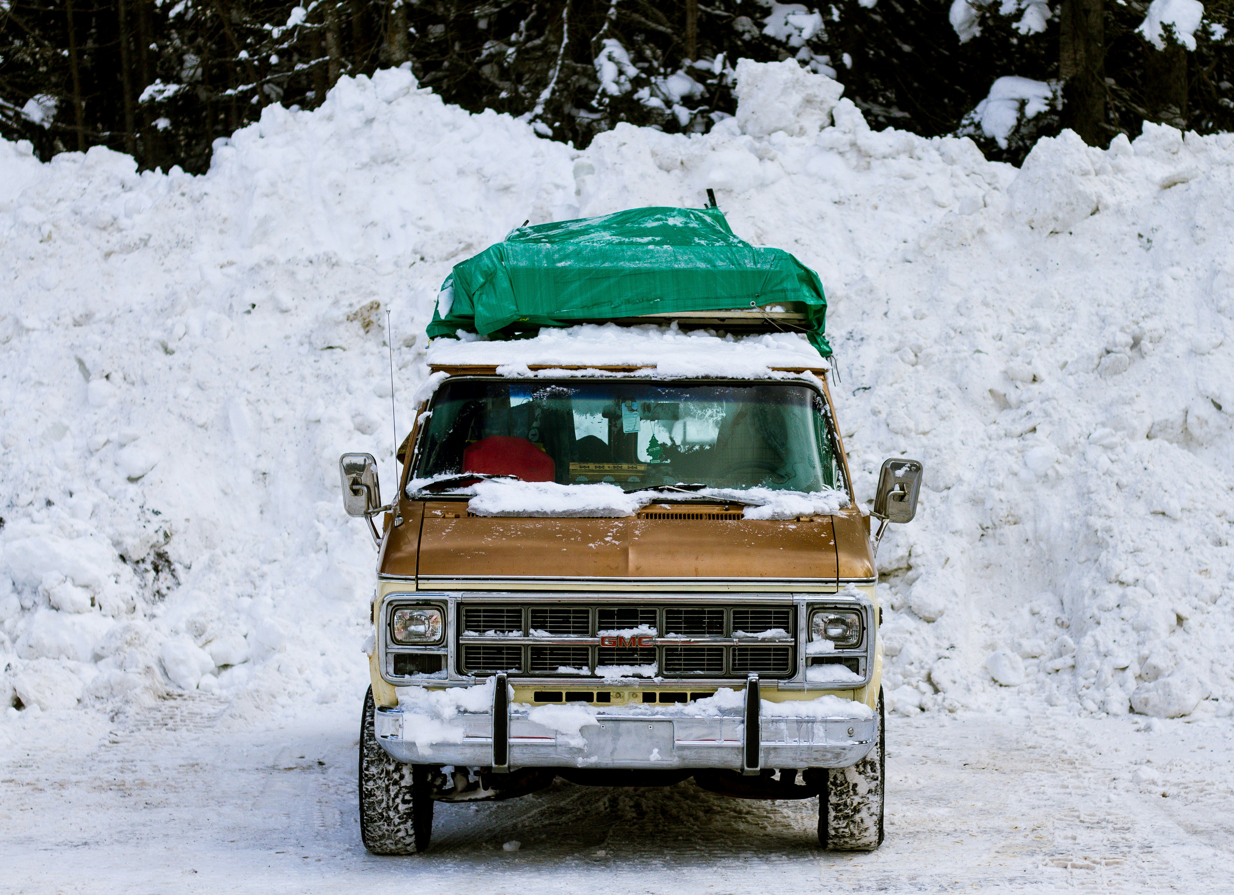 A vintage van, partially buried in snow, with a green tarp atop, set against a backdrop of snow-laden trees. The scene conveys a sense of isolation and adventure.