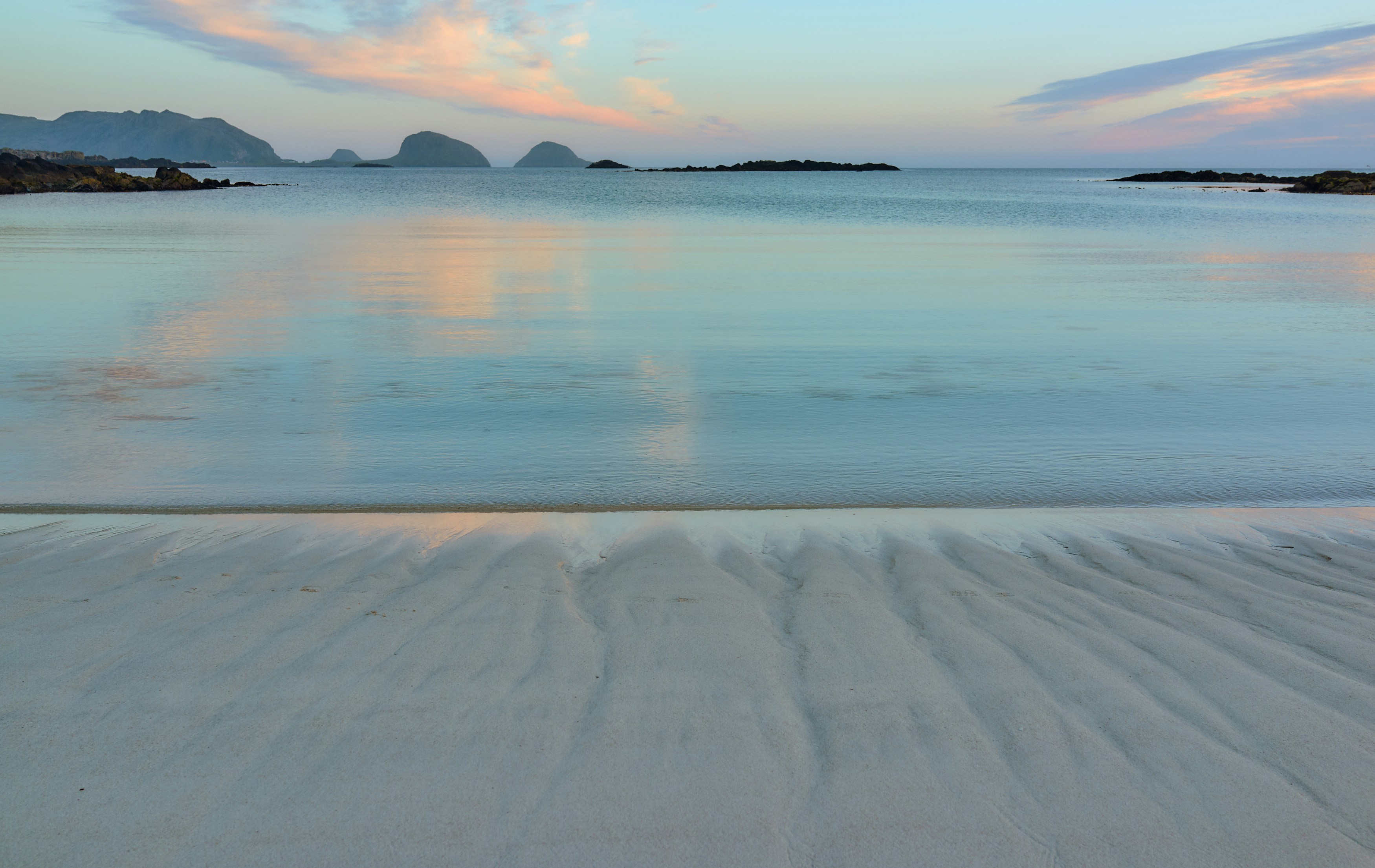 Gentle waves lap against a smooth sandy beach, with distant islands silhouetted against a pastel sky at twilight.