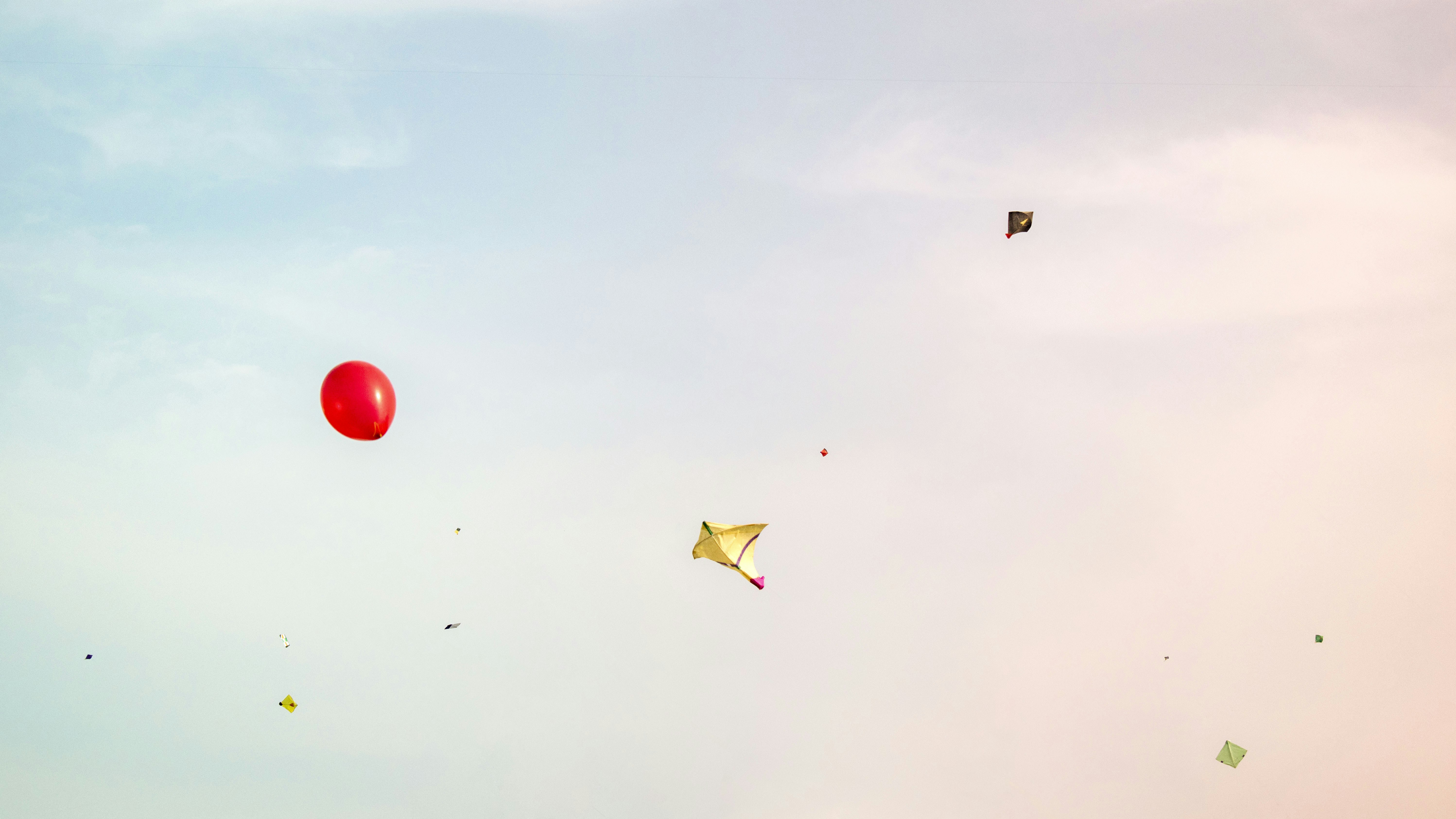 Colorful kites and a bright red balloon float against a soft pastel sky, evoking a sense of freedom and joy.