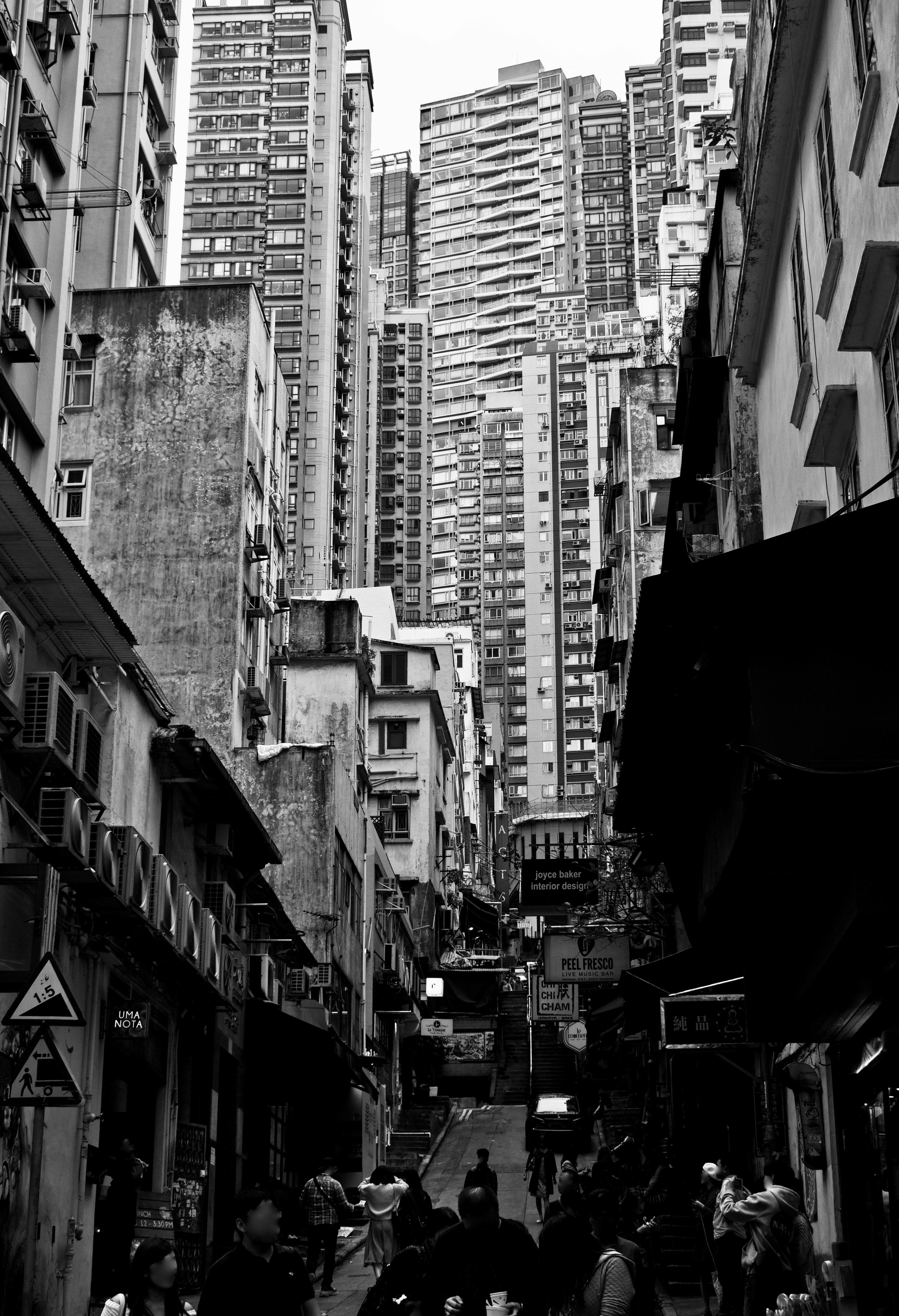 Narrow street lined with towering high-rise buildings in monochrome, showcasing the contrast between urban life and architecture. People navigate the bustling scene.