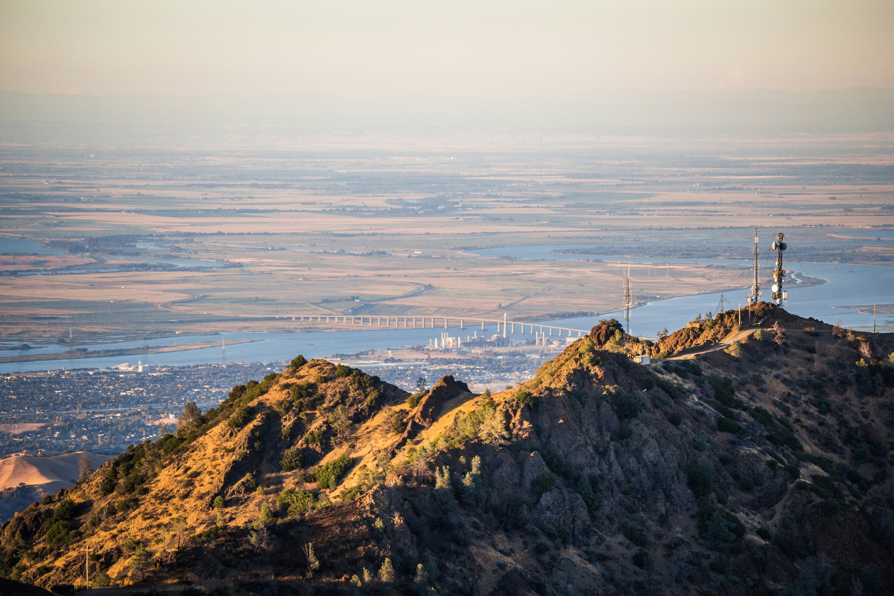 Mountain peaks with communication towers overlooking a cityscape and river at sunset.