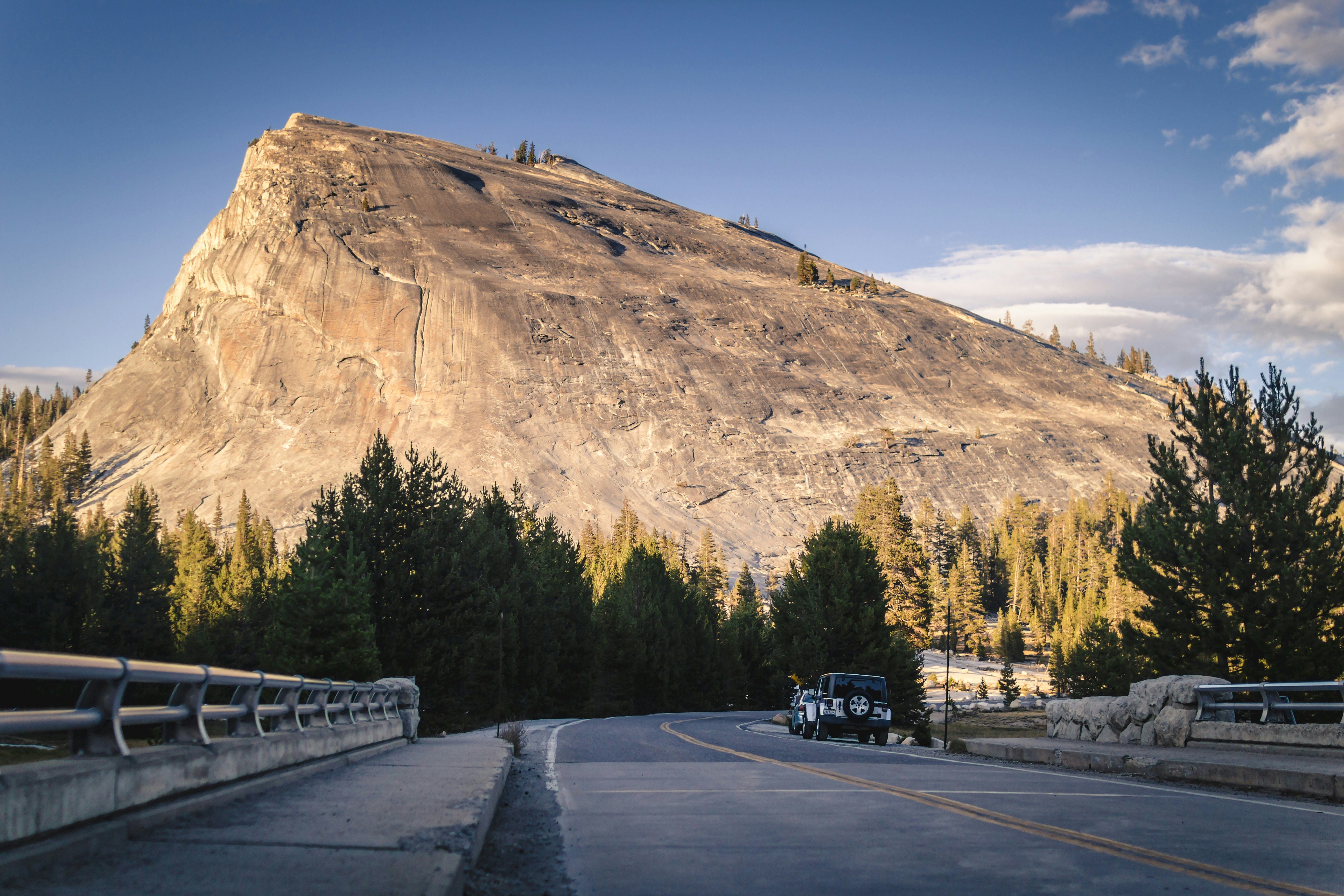 Massive granite rock formation rises above a winding road, framed by lush pine trees under a clear sky.