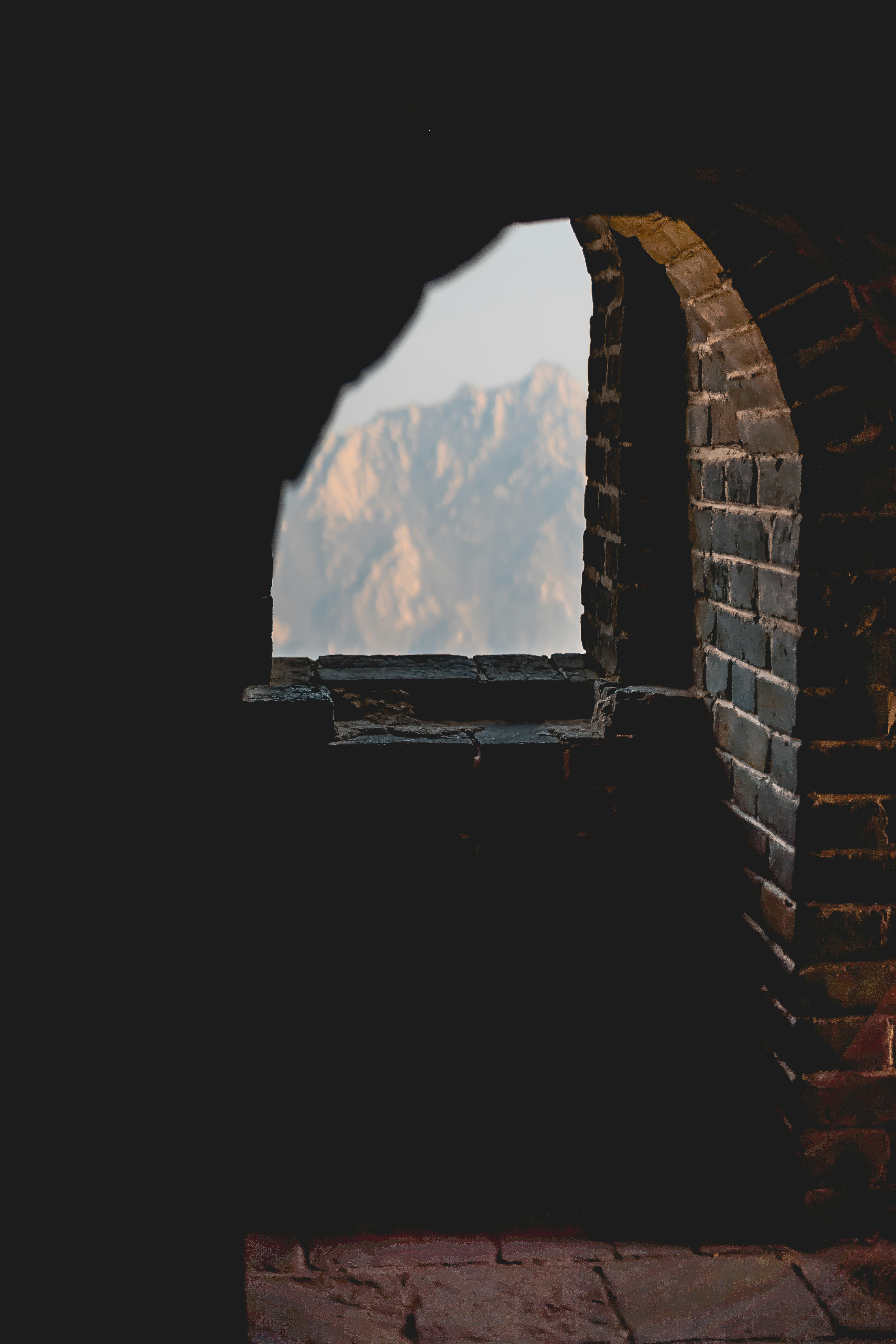 View through a stone archway showcasing distant mountains bathed in soft light. The historical architecture contrasts with the natural landscape.