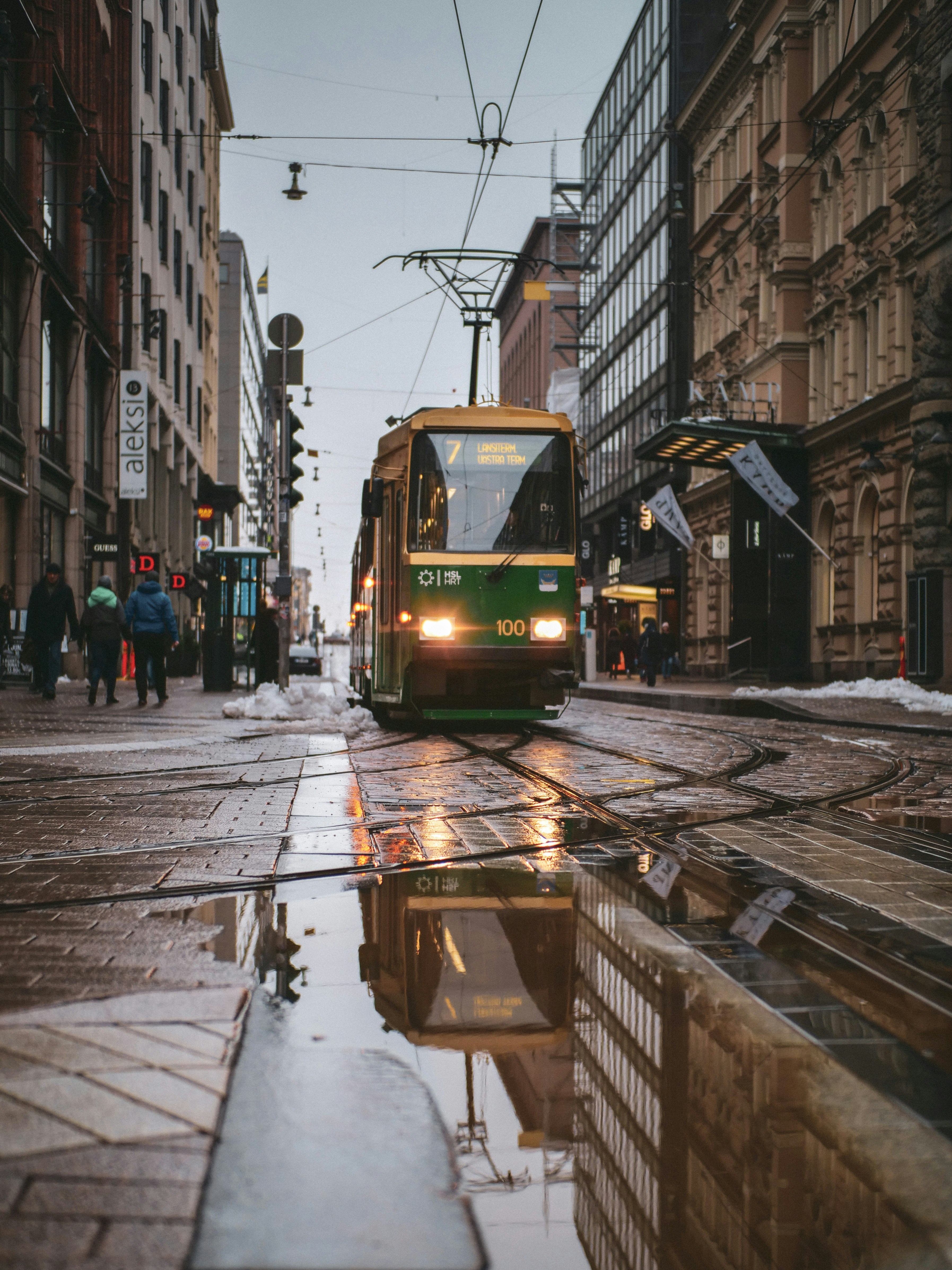 Green tram navigating a city street, reflecting in puddles on the pavement. People stroll along the sidewalk, surrounded by urban architecture.
