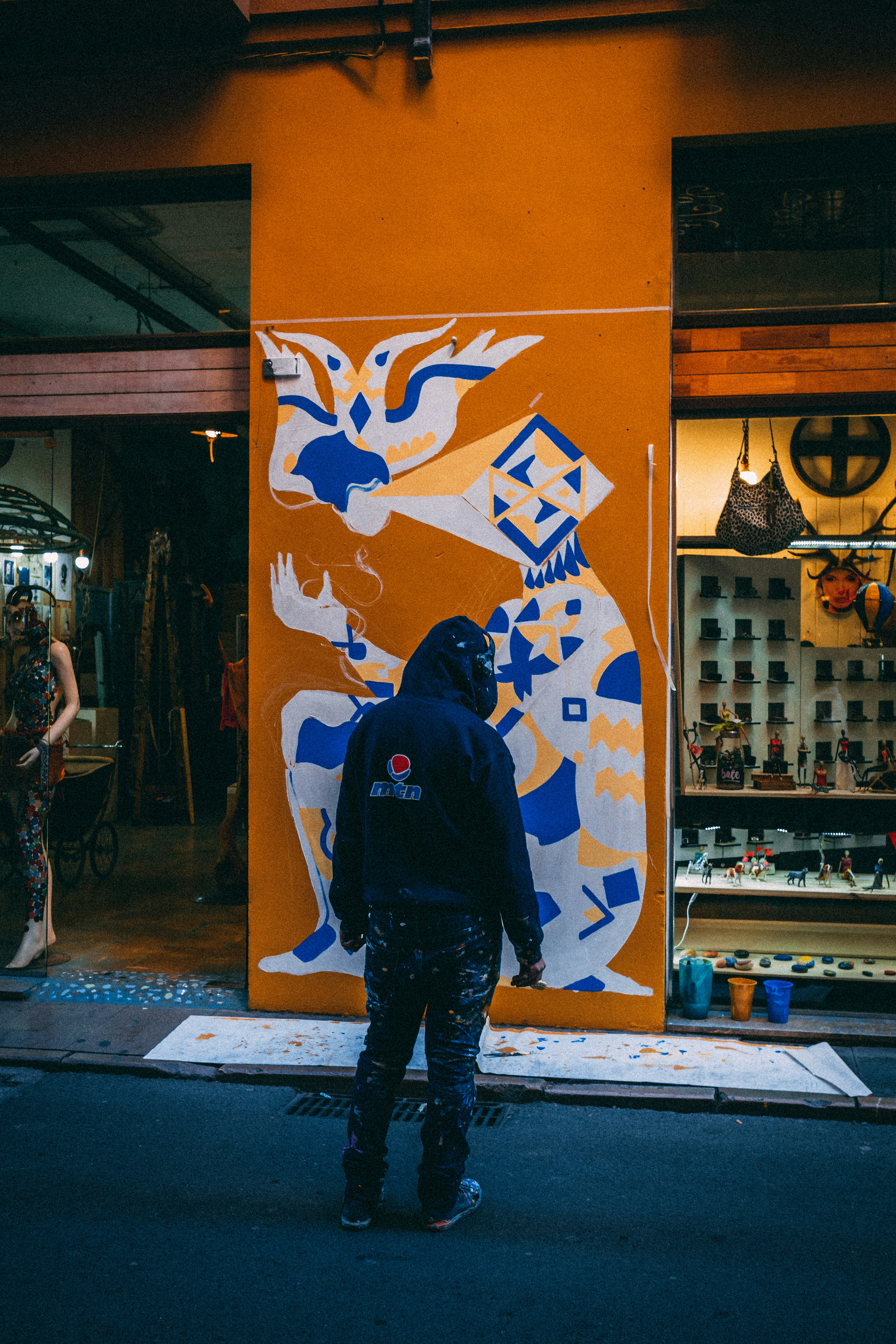 A figure in a dark hoodie stands before a vibrant mural on an orange wall, showcasing abstract shapes and forms. The scene captures the intersection of street art and urban life.