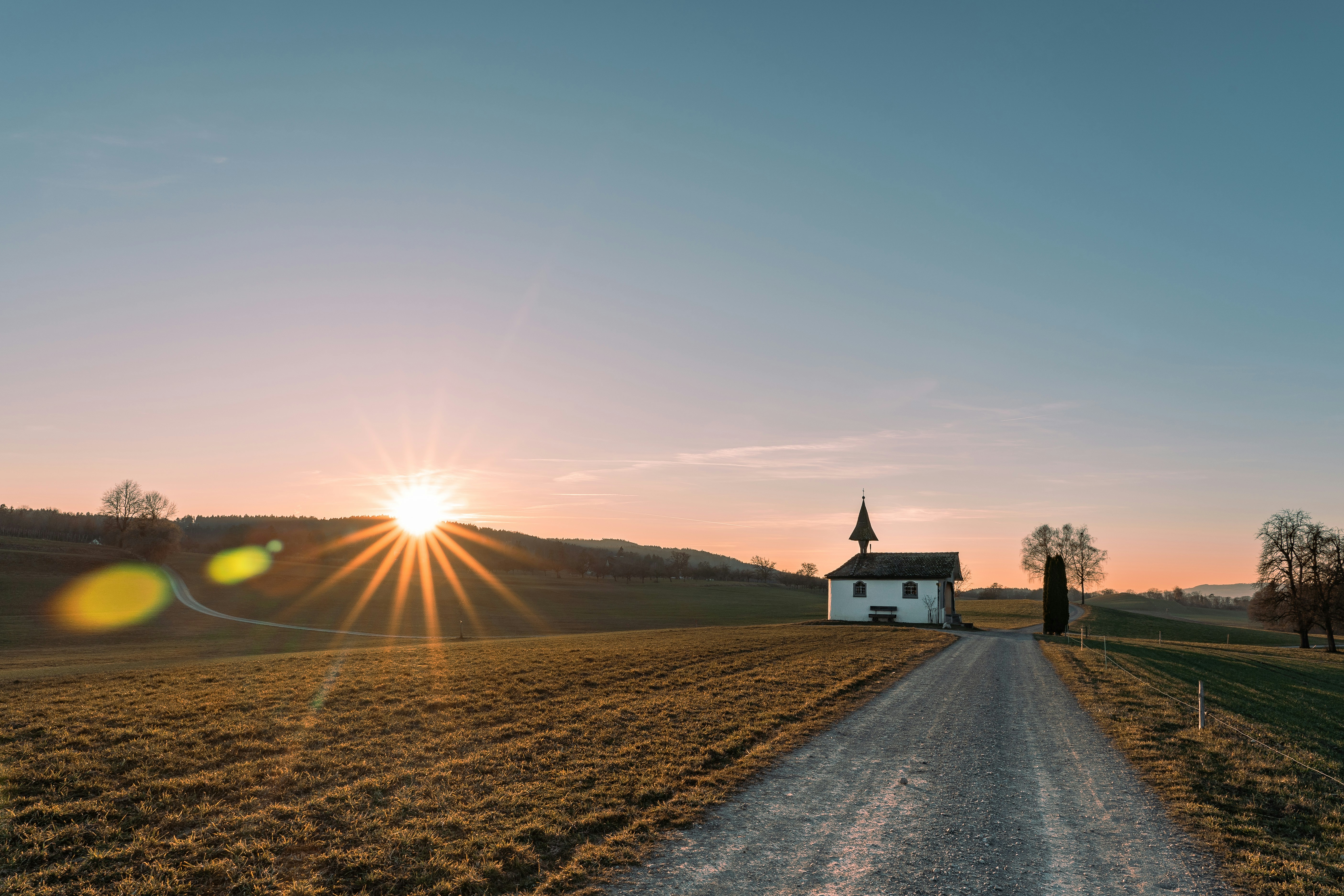 A quaint chapel stands beside a gravel path as the sun rises, casting rays across the tranquil countryside. The scene captures the peaceful transition from night to day.