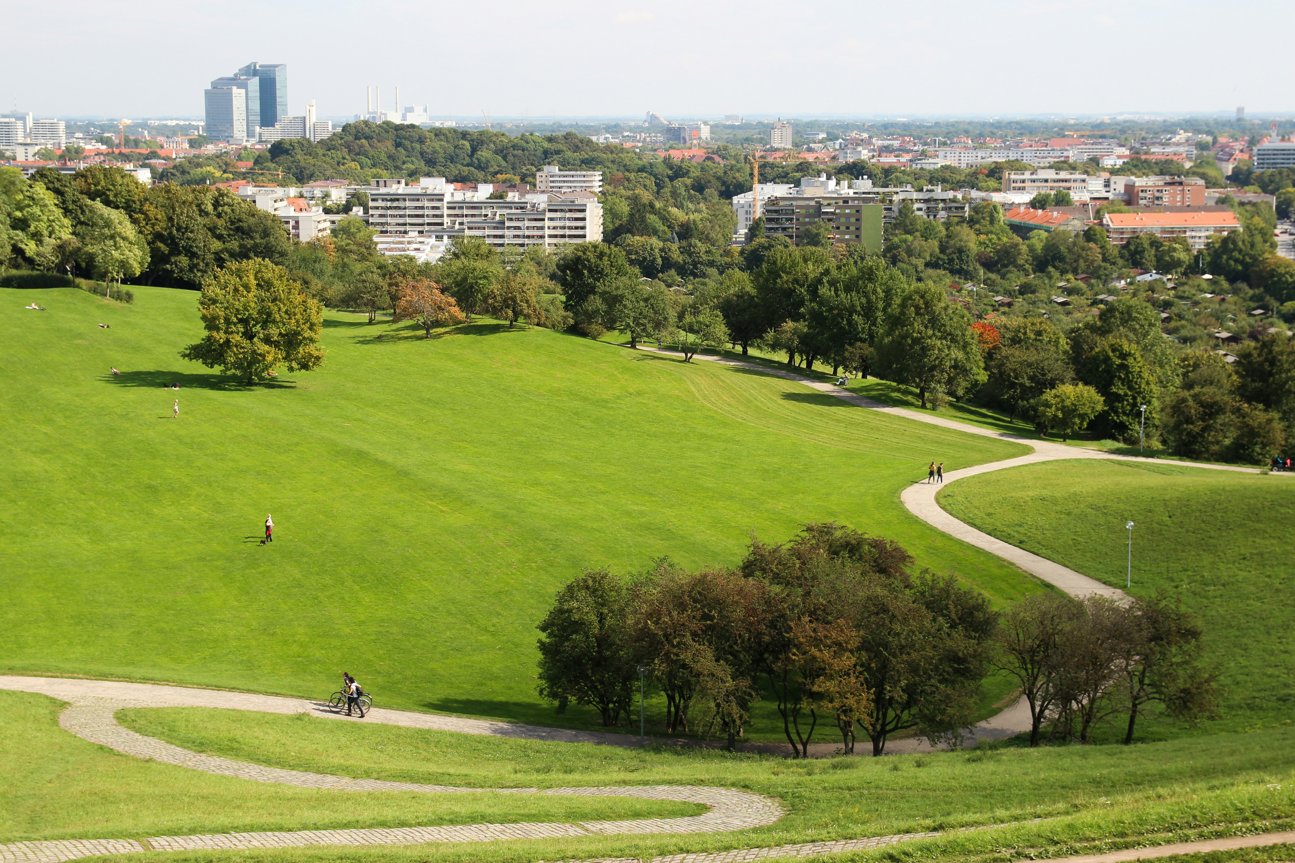 Curving paths wind through a lush green park with trees, set against a distant urban skyline.