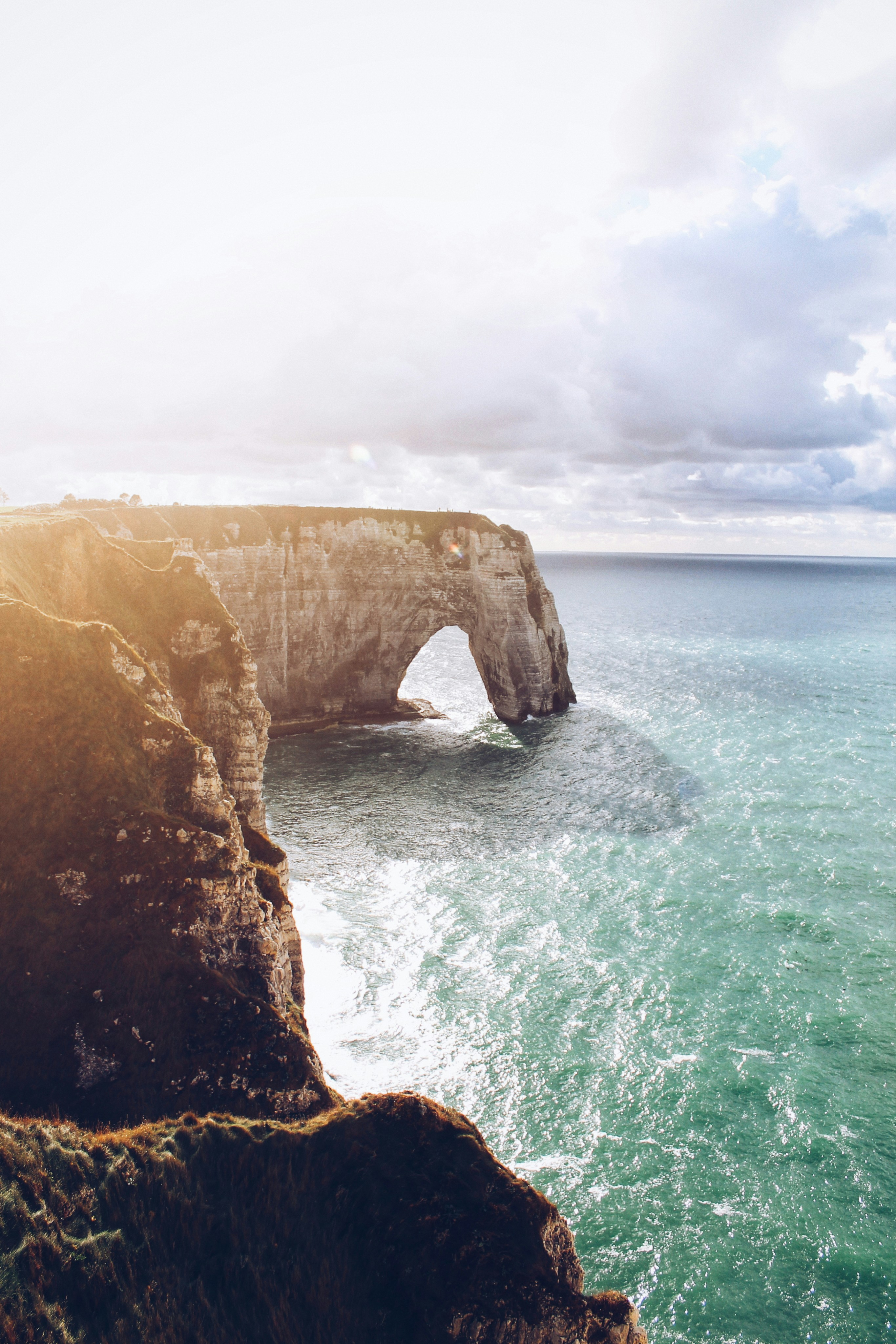 Dramatic coastal arch rising from turquoise waters, framed by rugged cliffs and dynamic cloud formations.