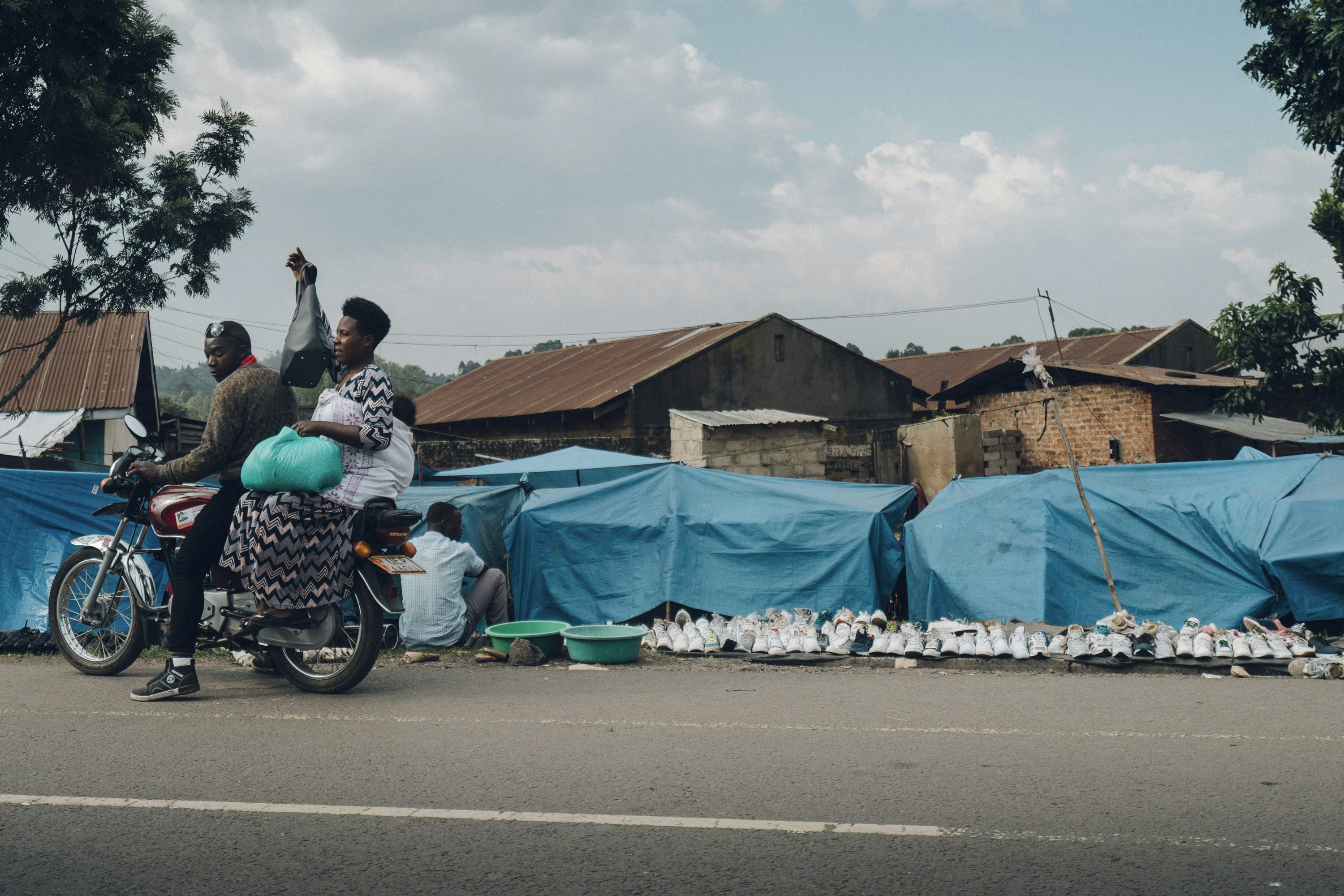 Motorcyclists ride past a vibrant street market with blue tarpaulins and rustic buildings under a cloudy sky.