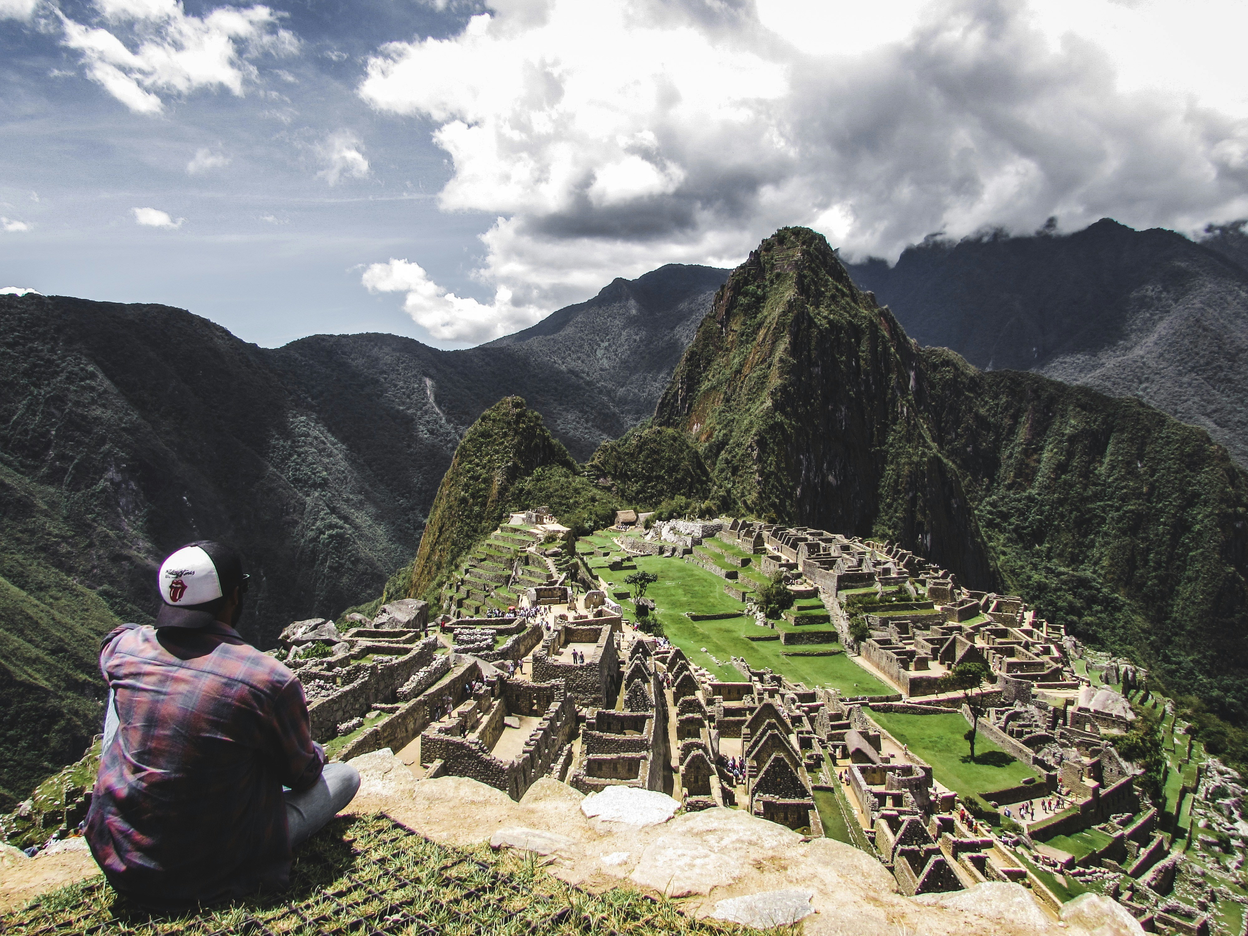 Person sitting on a hill overlooking the ancient site of Machu Picchu under a partly cloudy sky.