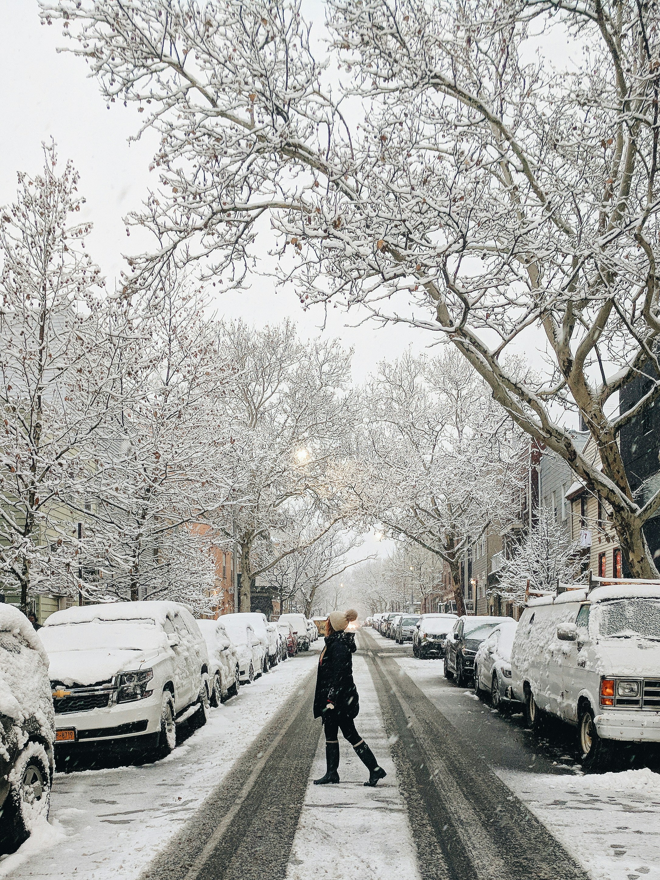 A solitary figure walks down a snow-covered street, flanked by frosted trees and parked cars blanketed in white. The soft glow of streetlights peeks through the winter haze.