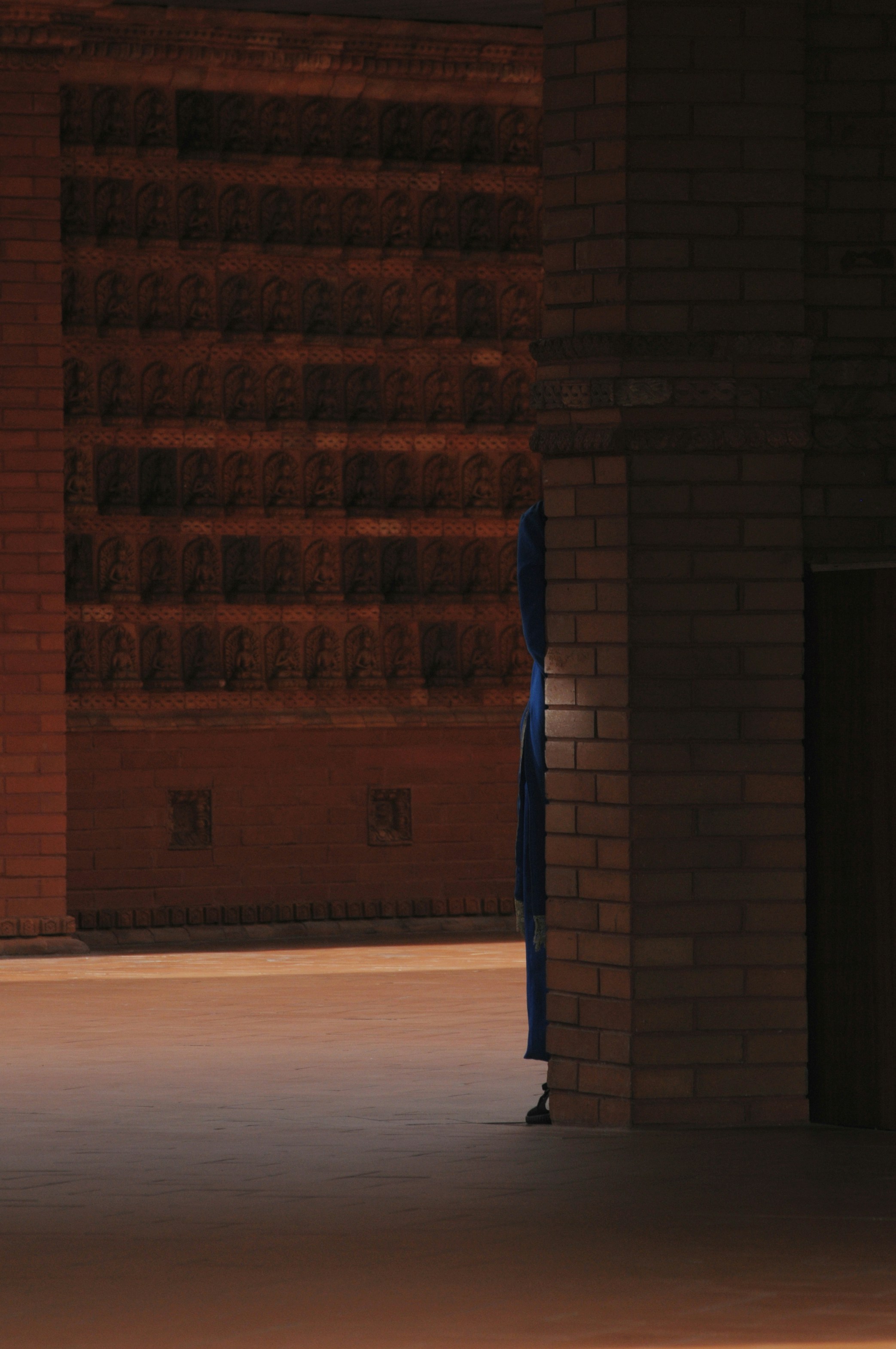 A serene interior showcasing intricate brickwork and sculptures, with a figure partially obscured in a blue garment. The interplay of light and shadow adds depth to the scene.