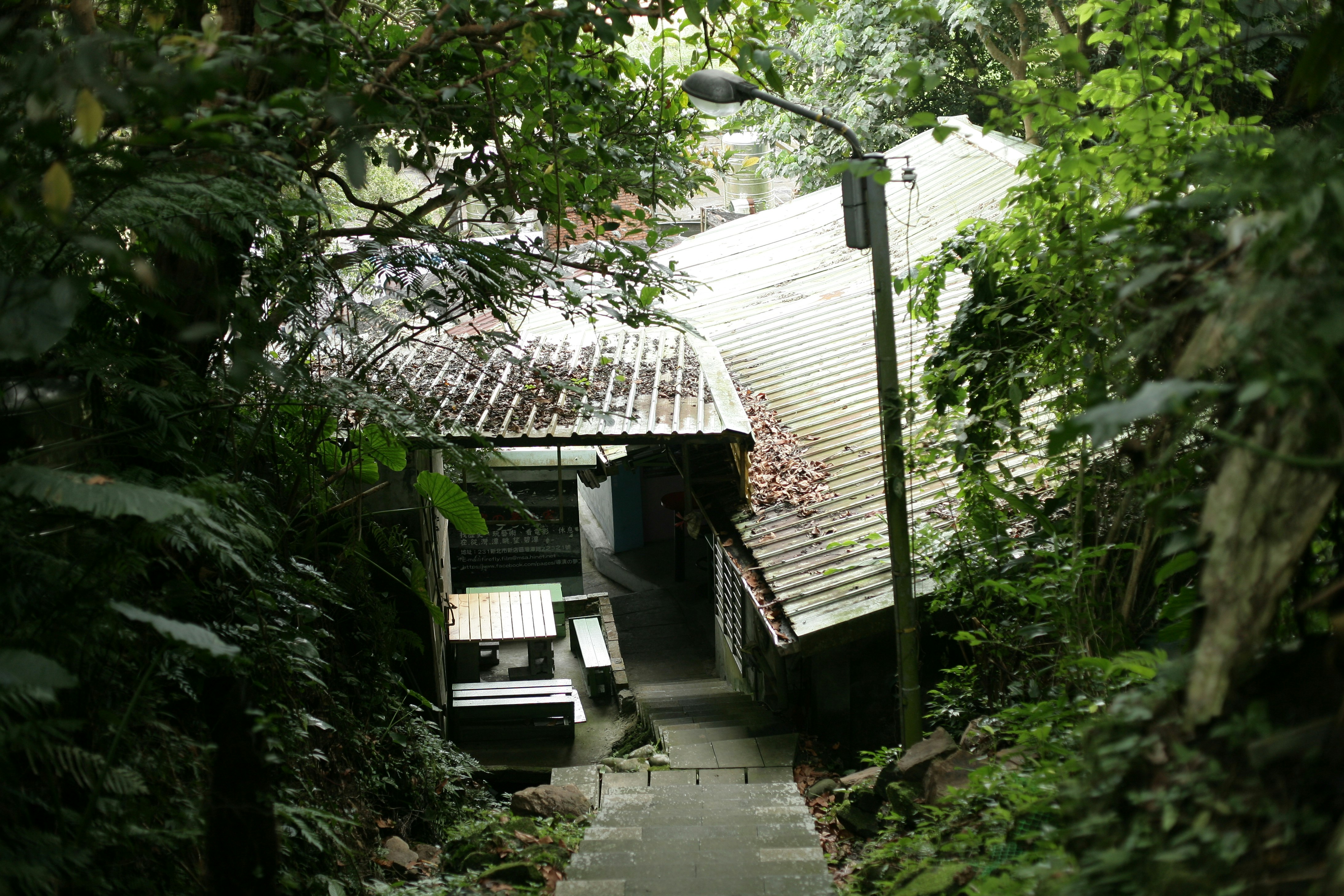 A rustic building nestled within lush greenery, accessible by a stone pathway surrounded by vibrant foliage. The scene evokes a sense of tranquility and seclusion.