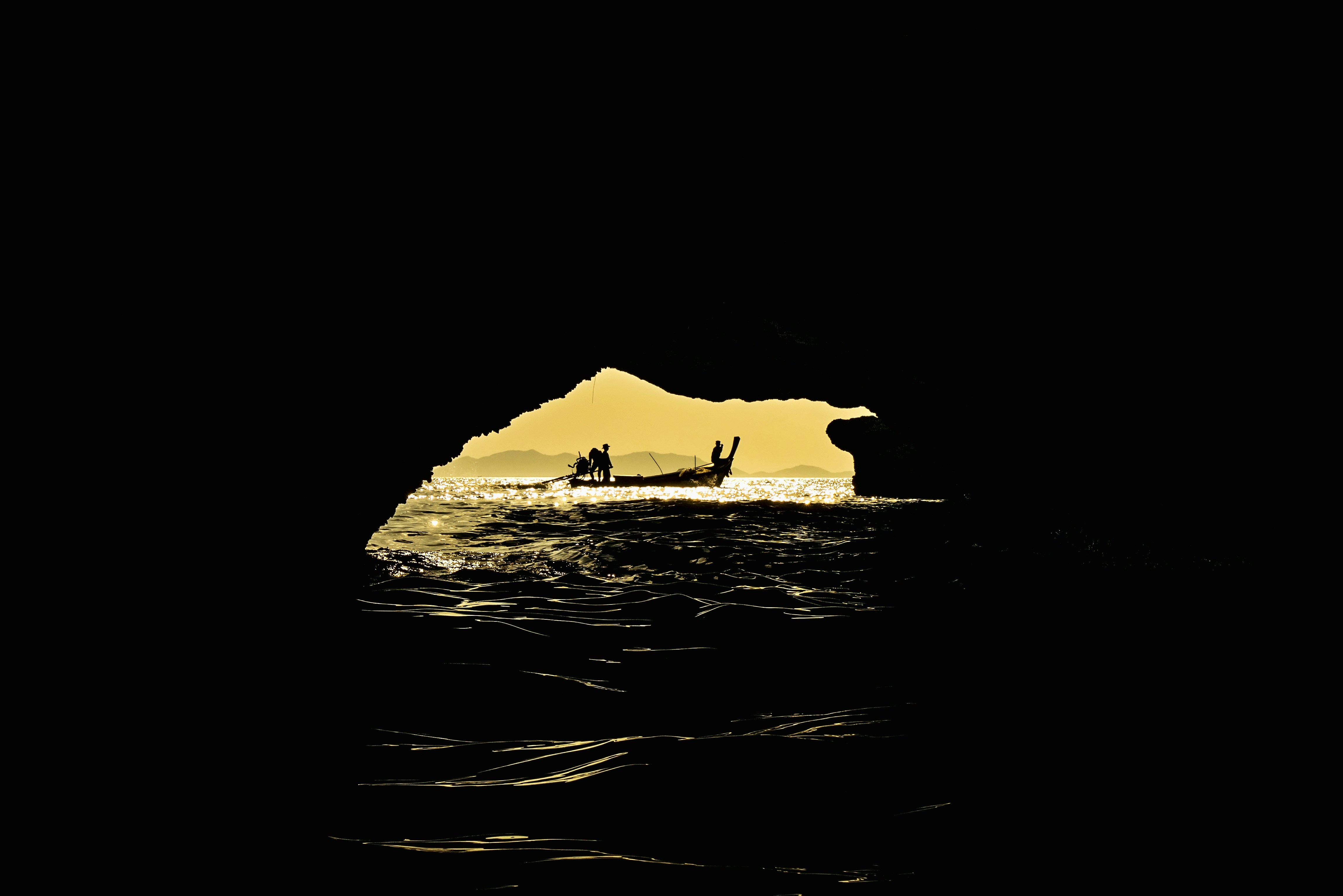Two paddlers are silhouetted as they pass through a sea-cave opening, with a warm sunset glow illuminating the horizon. The scene emphasizes the contrast between dark foreground and the bright water and sky.